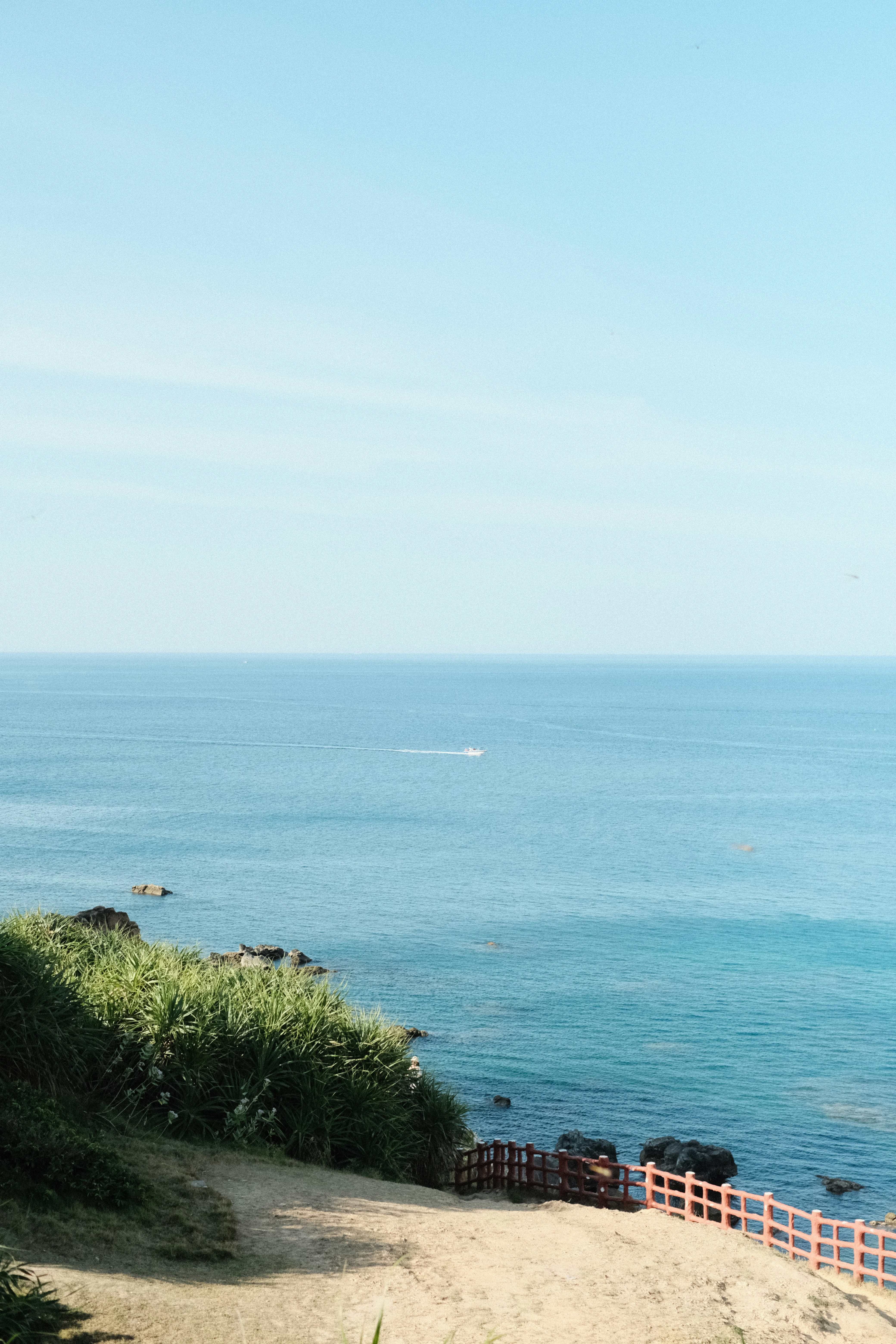 A tranquil coastal scene featuring a distant boat navigating the calm waters, framed by lush green foliage and a wooden railing. The clear blue sky enhances the serene atmosphere.