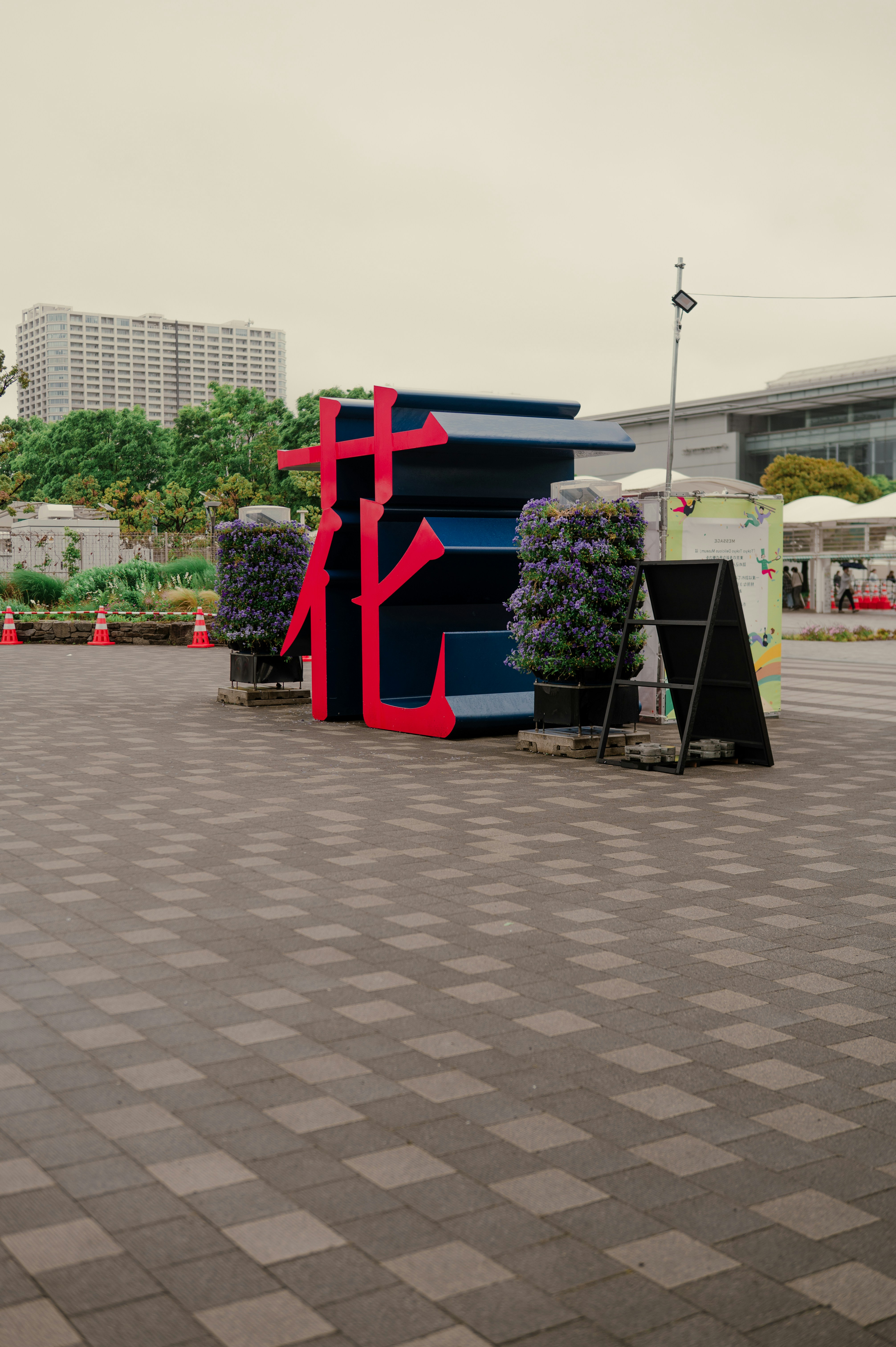 A vibrant red floral-themed installation stands amidst a landscaped area, surrounded by lush greenery and decorative planters. The scene captures a blend of nature and contemporary design.