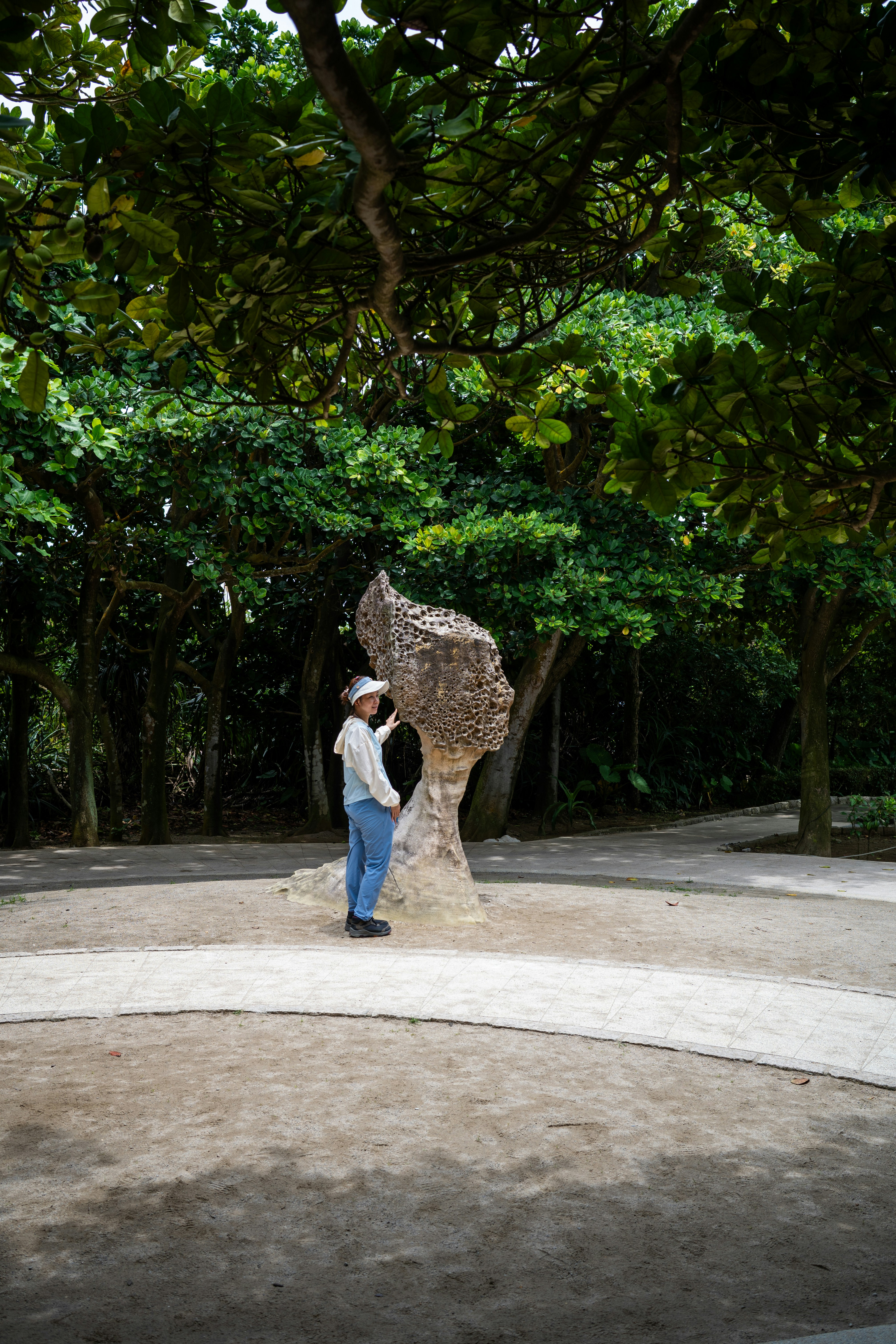 A person stands beside a unique rock formation in a lush green park, surrounded by trees. The scene captures a moment of contemplation amid nature's artistry.