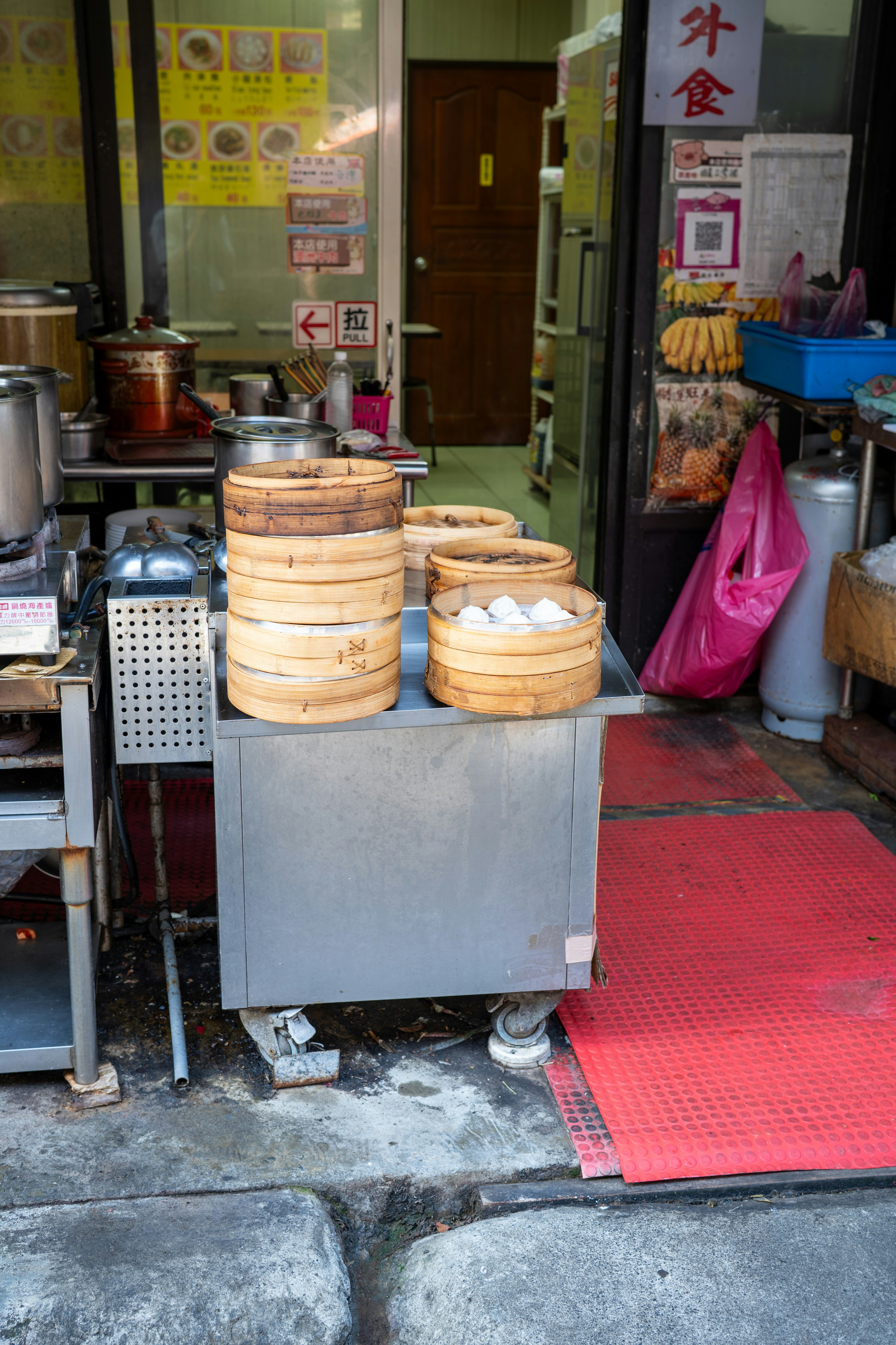 Bamboo steamers filled with dumplings rest on a metal cart outside a bustling food stall, showcasing the vibrant street food scene.