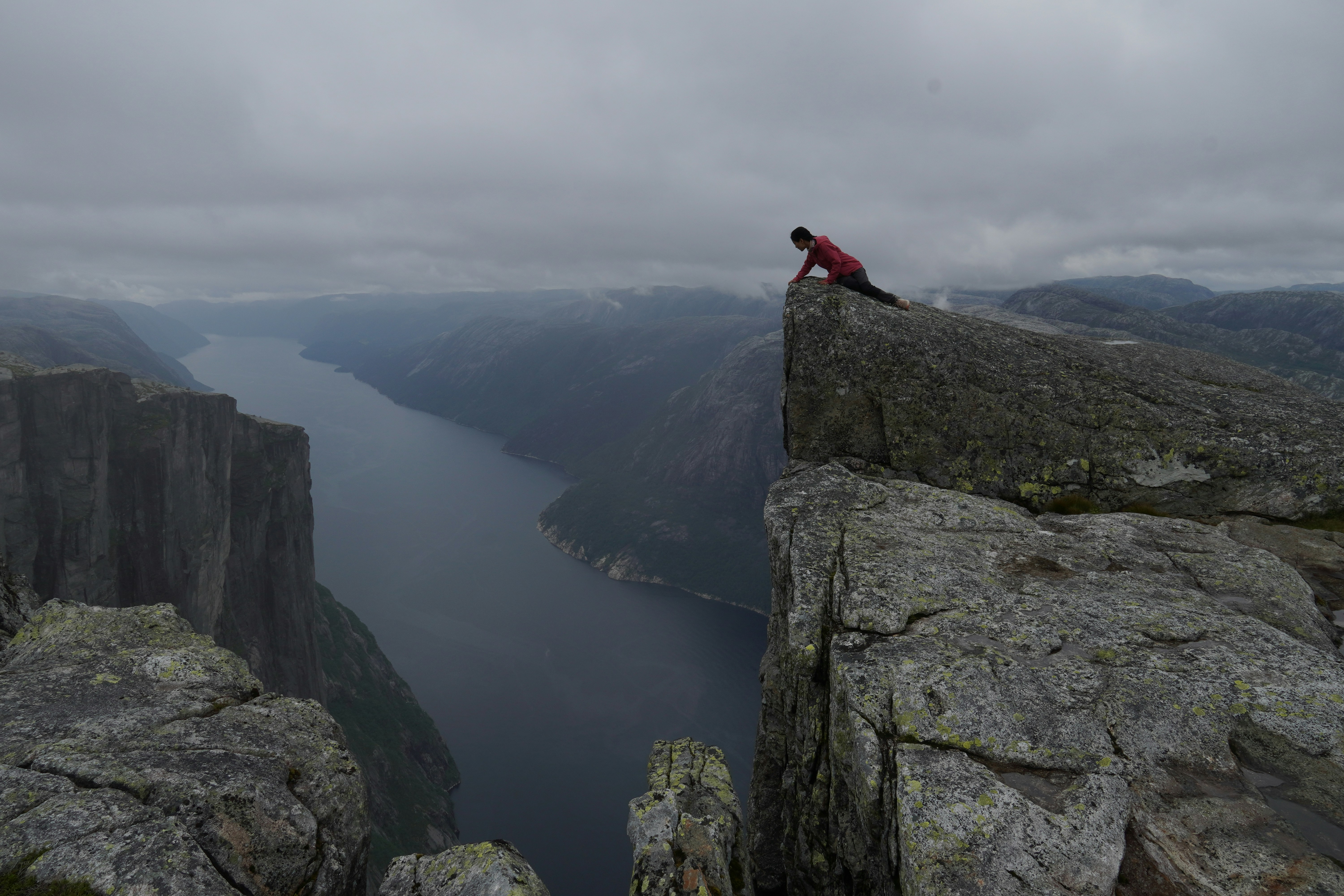 Climber leaning over a rocky precipice, gazing at the expansive fjord below, surrounded by dramatic cliffs and overcast skies.
