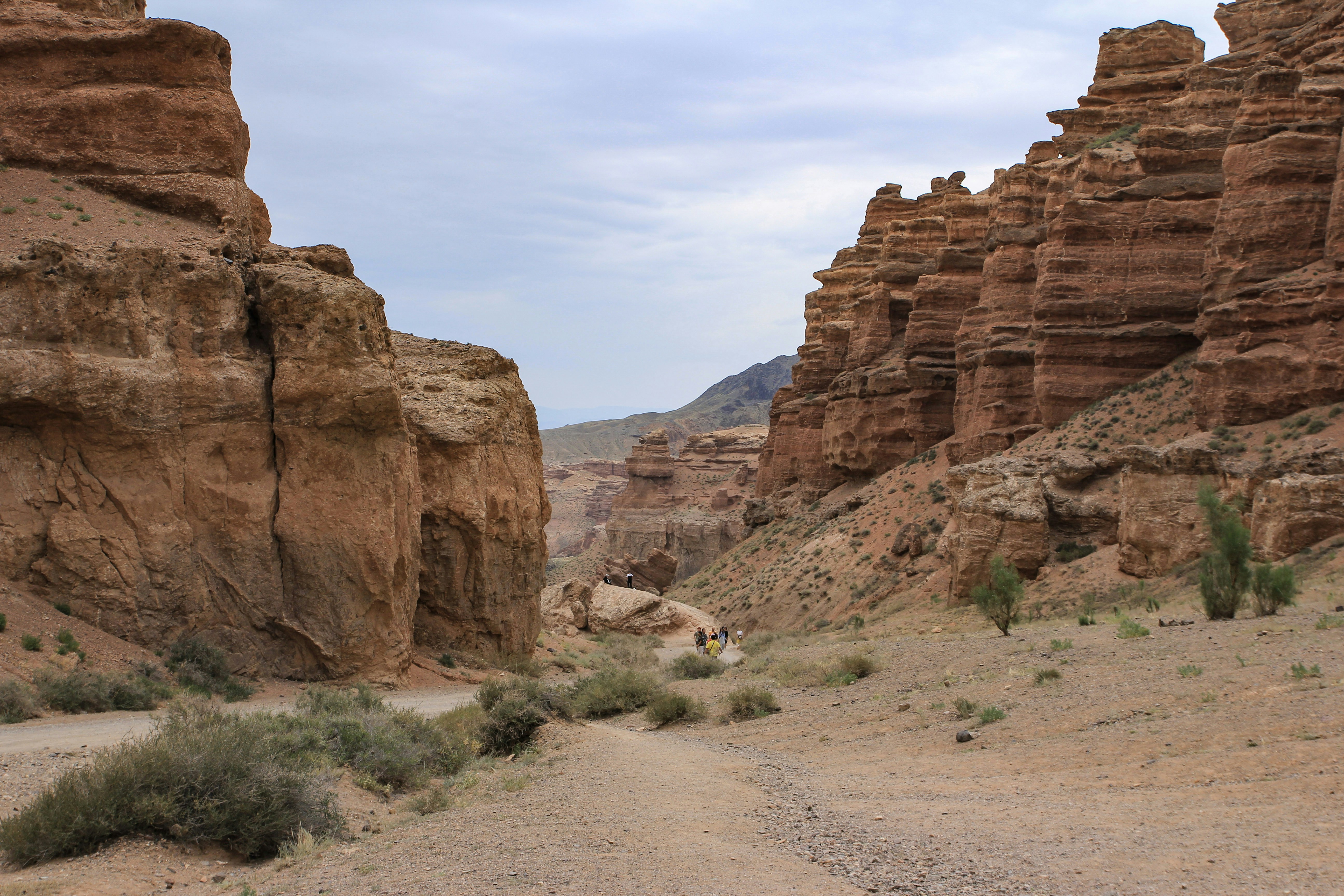 Winding path through rugged canyon landscape, flanked by towering red rock formations. A hint of distant figures adds scale to the vastness.