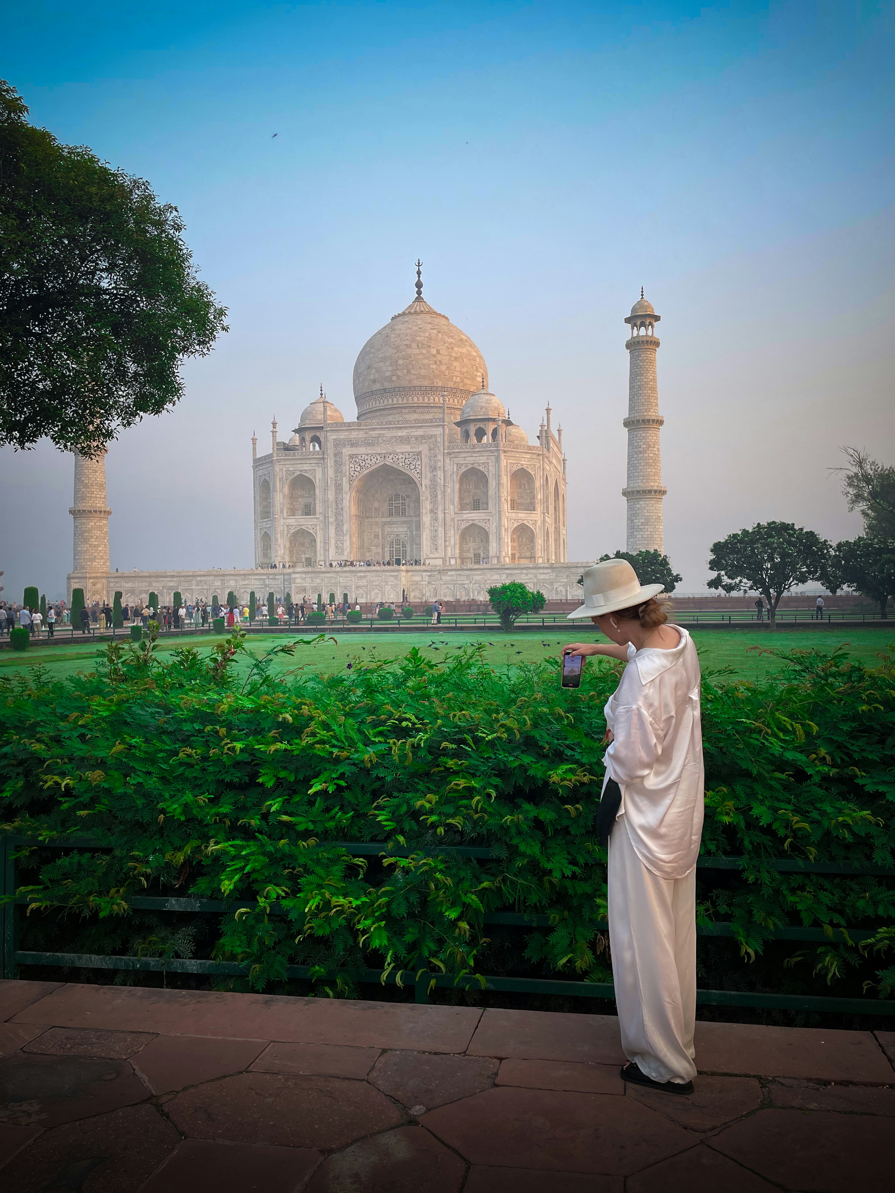 A woman dressed in white stands gracefully in front of the Taj Mahal, capturing its grandeur on her phone. Lush greenery frames the iconic monument.