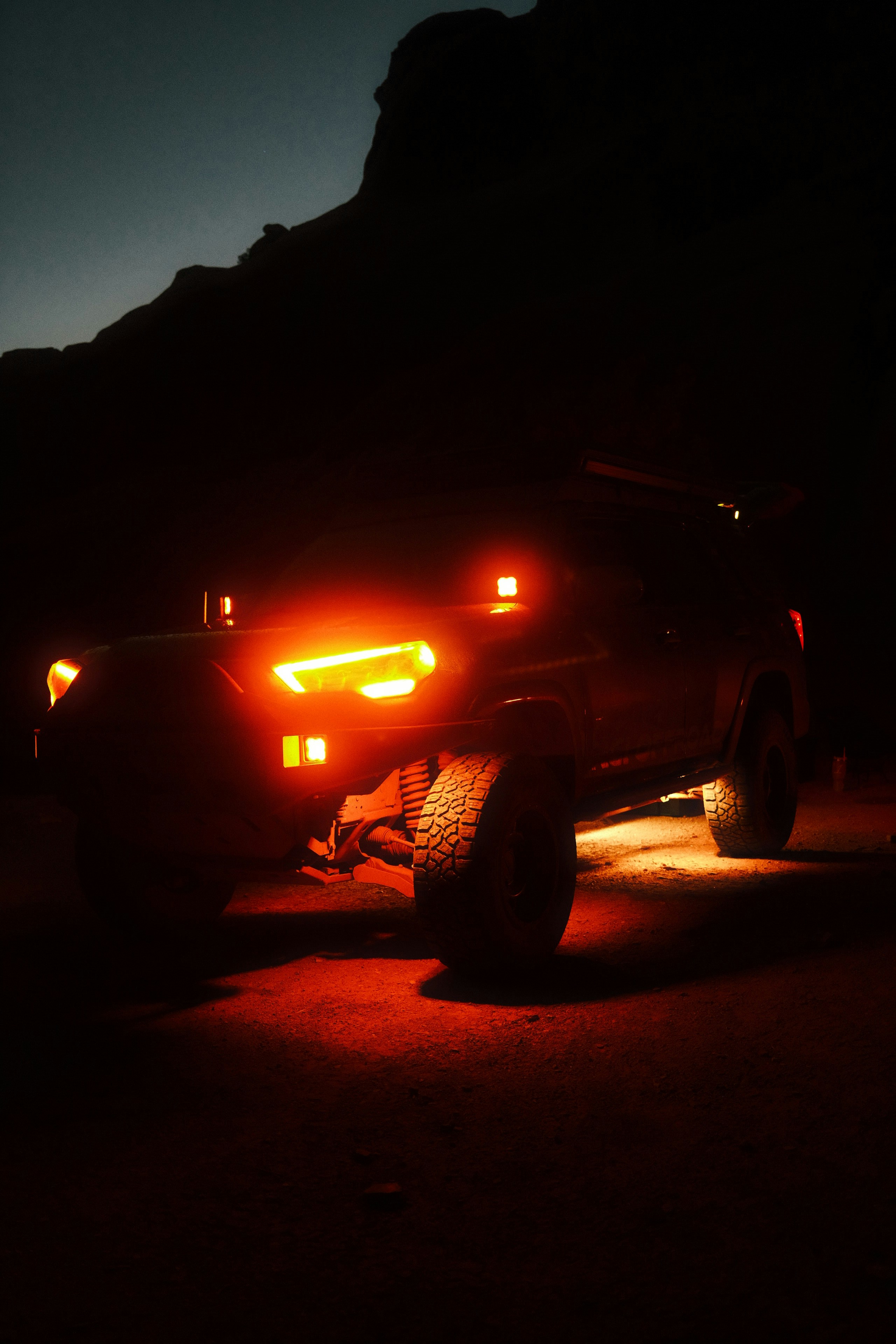 Off-road vehicle with vibrant orange lights glowing against a dark backdrop of rocky terrain.