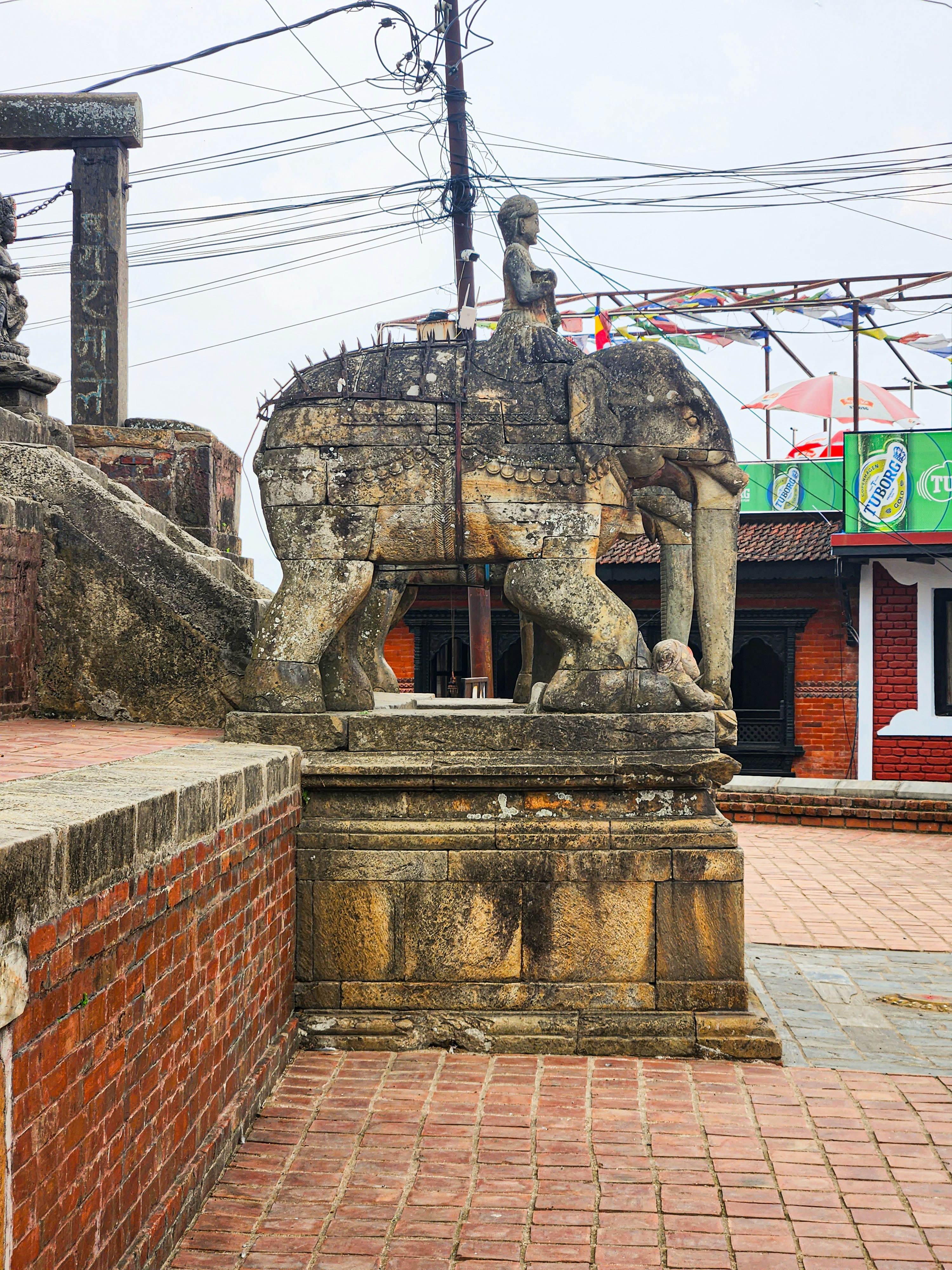 Ancient stone elephant statue with a seated figure atop, surrounded by traditional architecture and colorful banners in the background.