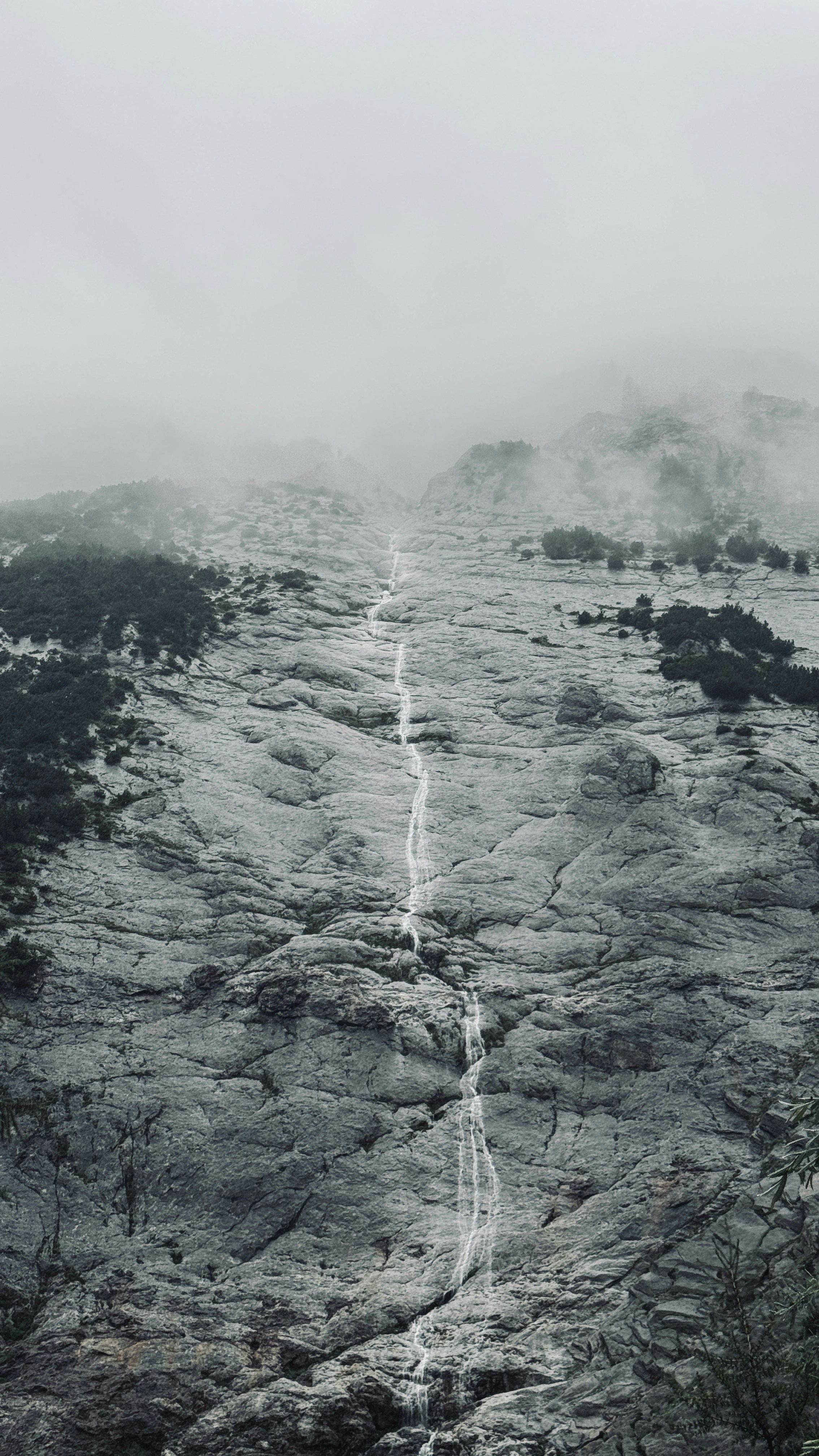 A cascading waterfall flows down a rocky mountain face, shrouded in mist and greenery. The scene captures the serene yet powerful essence of nature's beauty.
