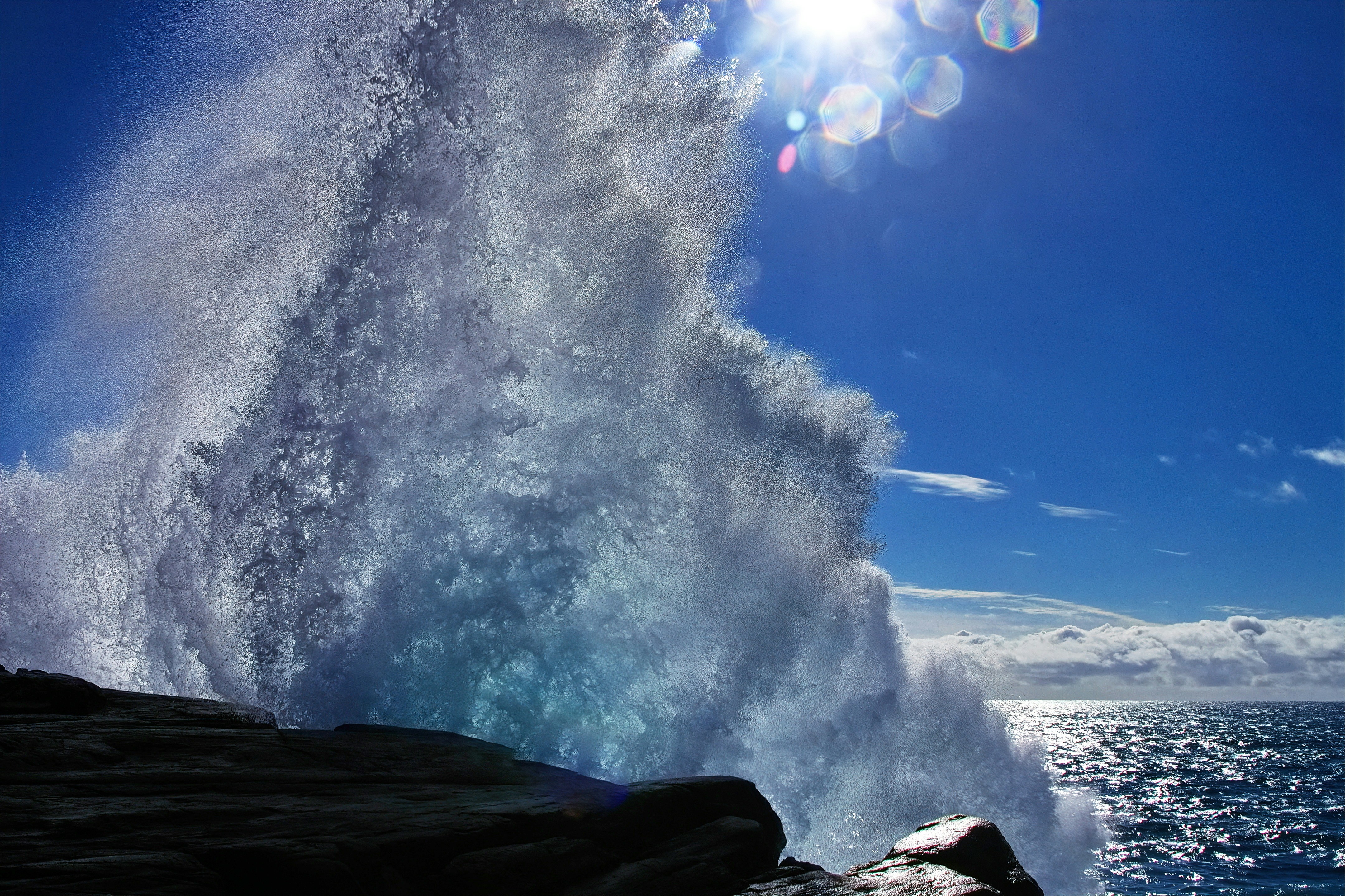 A powerful wave crashing against rocky shore under a bright blue sky, showcasing the dynamic interplay of water and light.