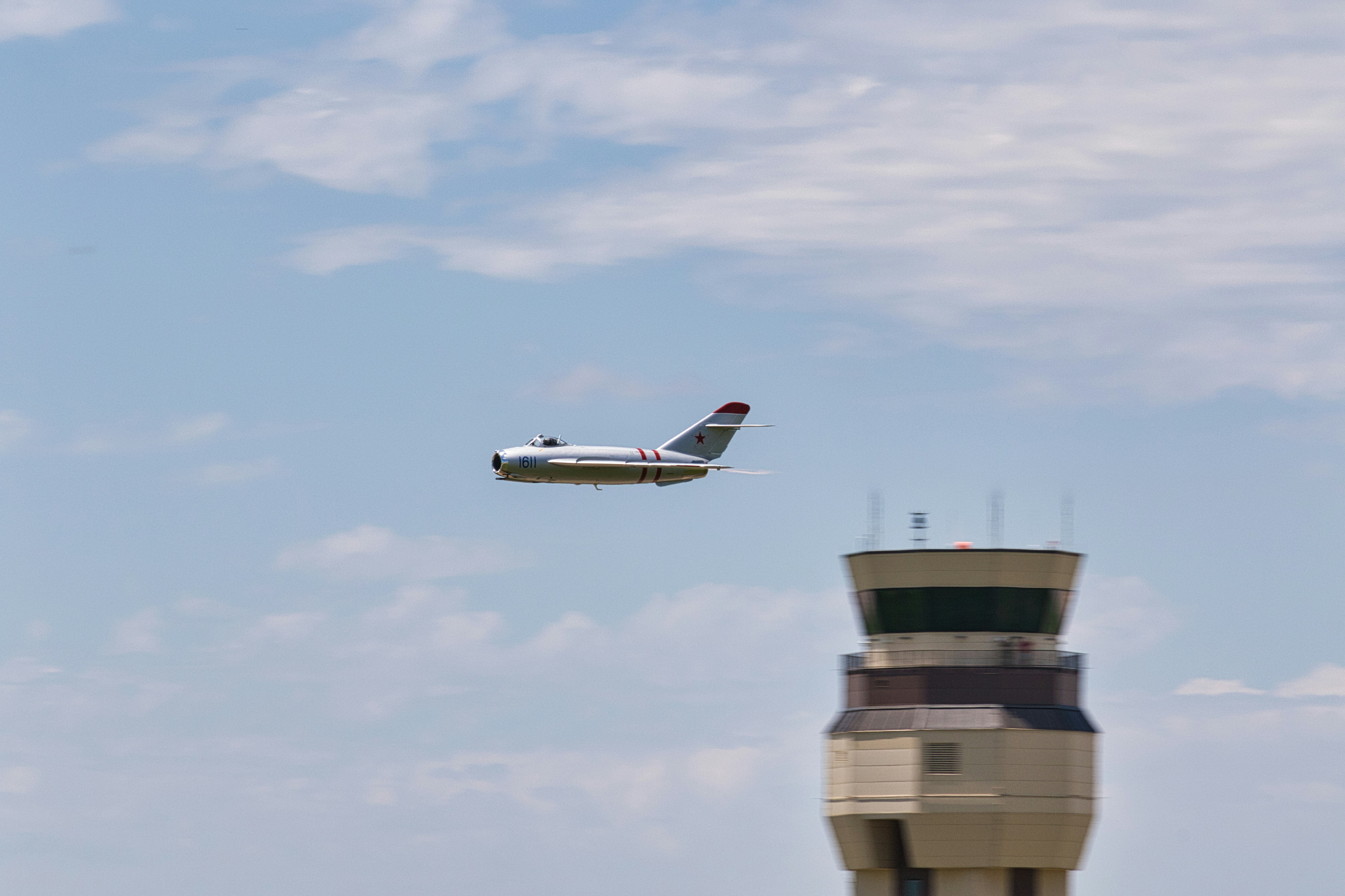 Business jet flying past an airport control tower under a bright sky.