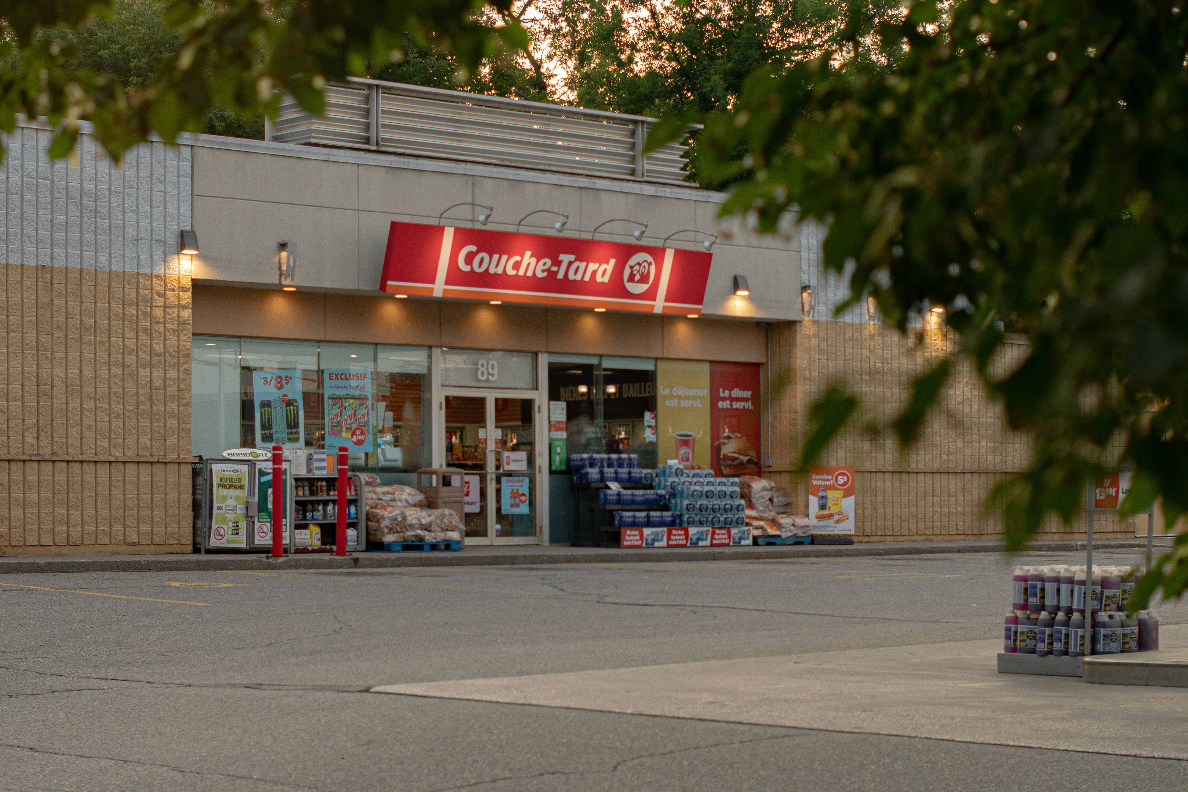 Convenience store entrance illuminated in the evening, showcasing a variety of products outside. The scene is framed by lush foliage.