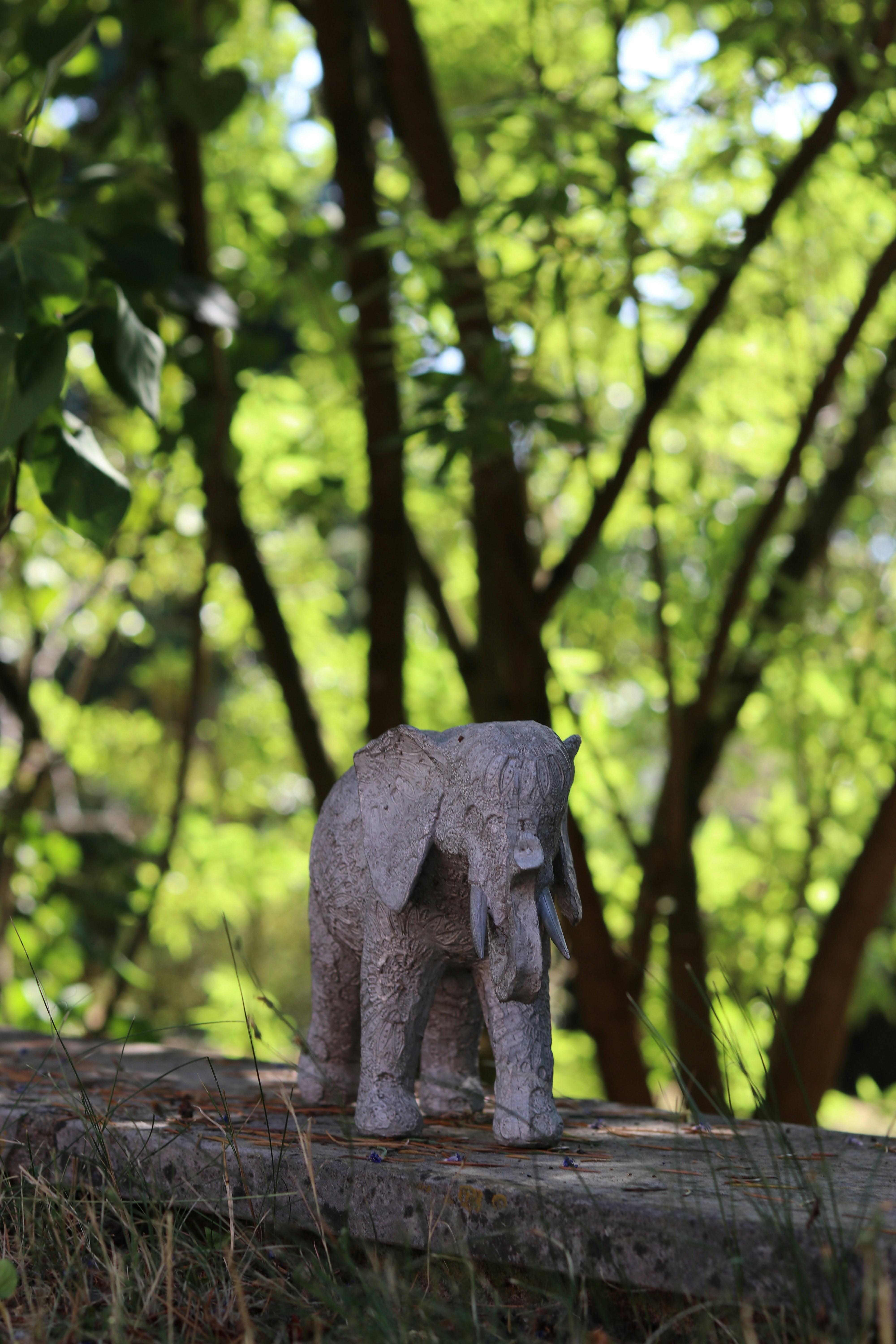 A textured stone elephant figurine stands atop a weathered slab, surrounded by tall grasses and framed by lush, sun‑dappled trees in a tranquil garden setting. | An elephant sculpture stands amid the trees.