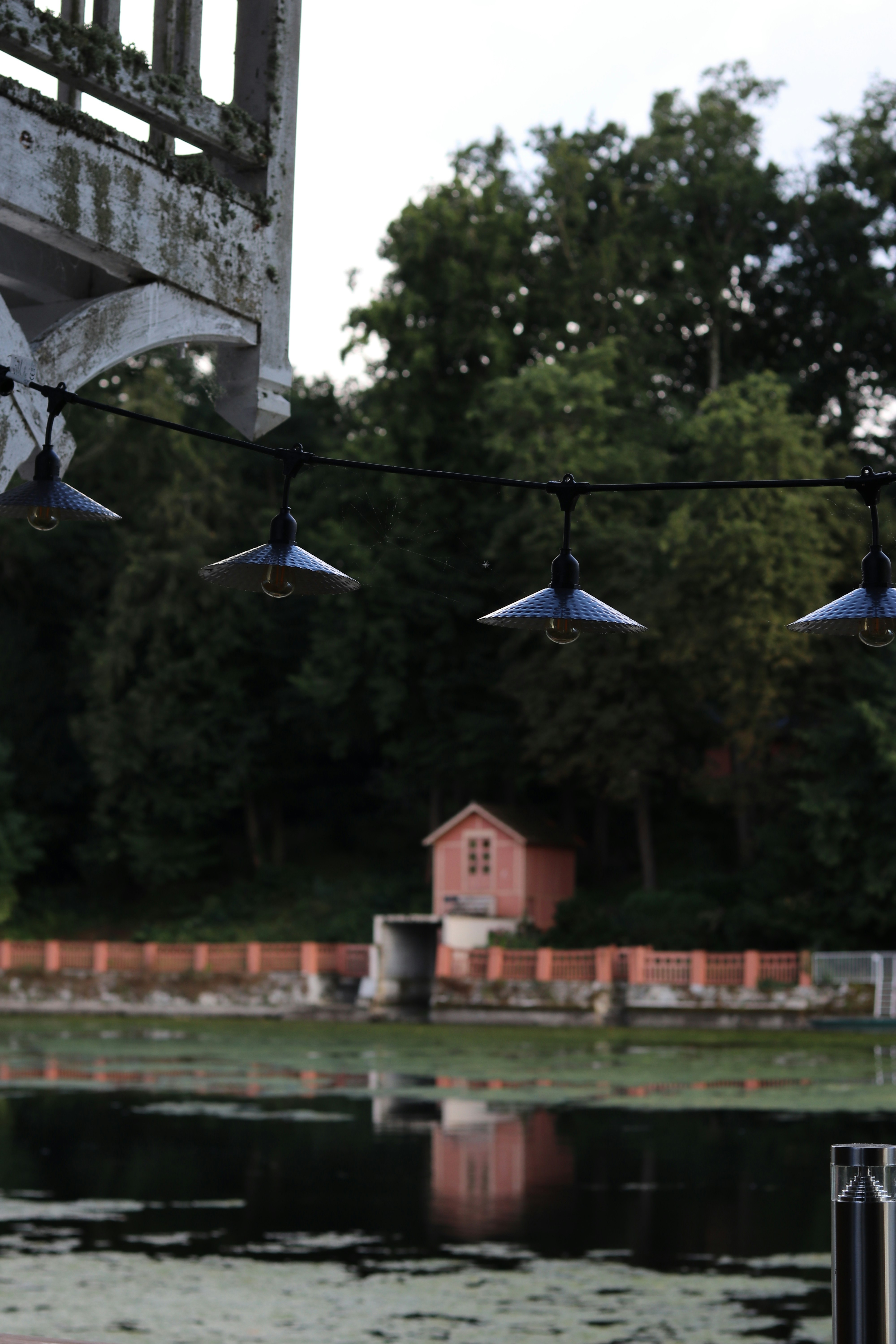 A row of metal pendant lights hangs beneath a weathered white balcony, framing a pastel pink boathouse and its reflection across a calm, algae‑dappled pond set against a lush forest backdrop. | String lights illuminate a lakefront scene at dusk.