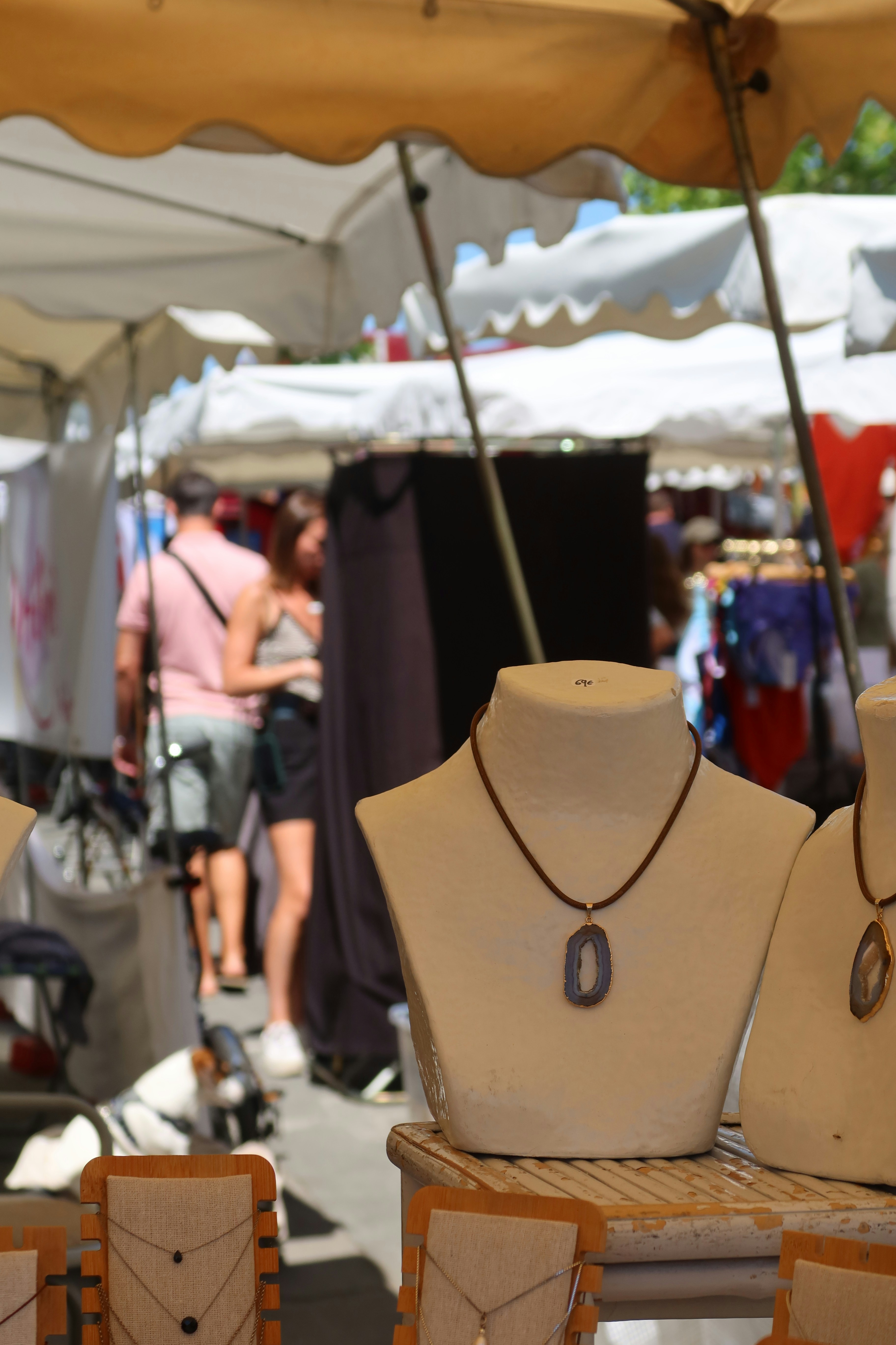 A geode slice pendant on a leather cord is showcased on a cream-colored bust at an outdoor market stall, with blurred tents, shoppers, and other jewelry displays creating a bustling summer bazaar backdrop. | Jewelry displayed at a market.