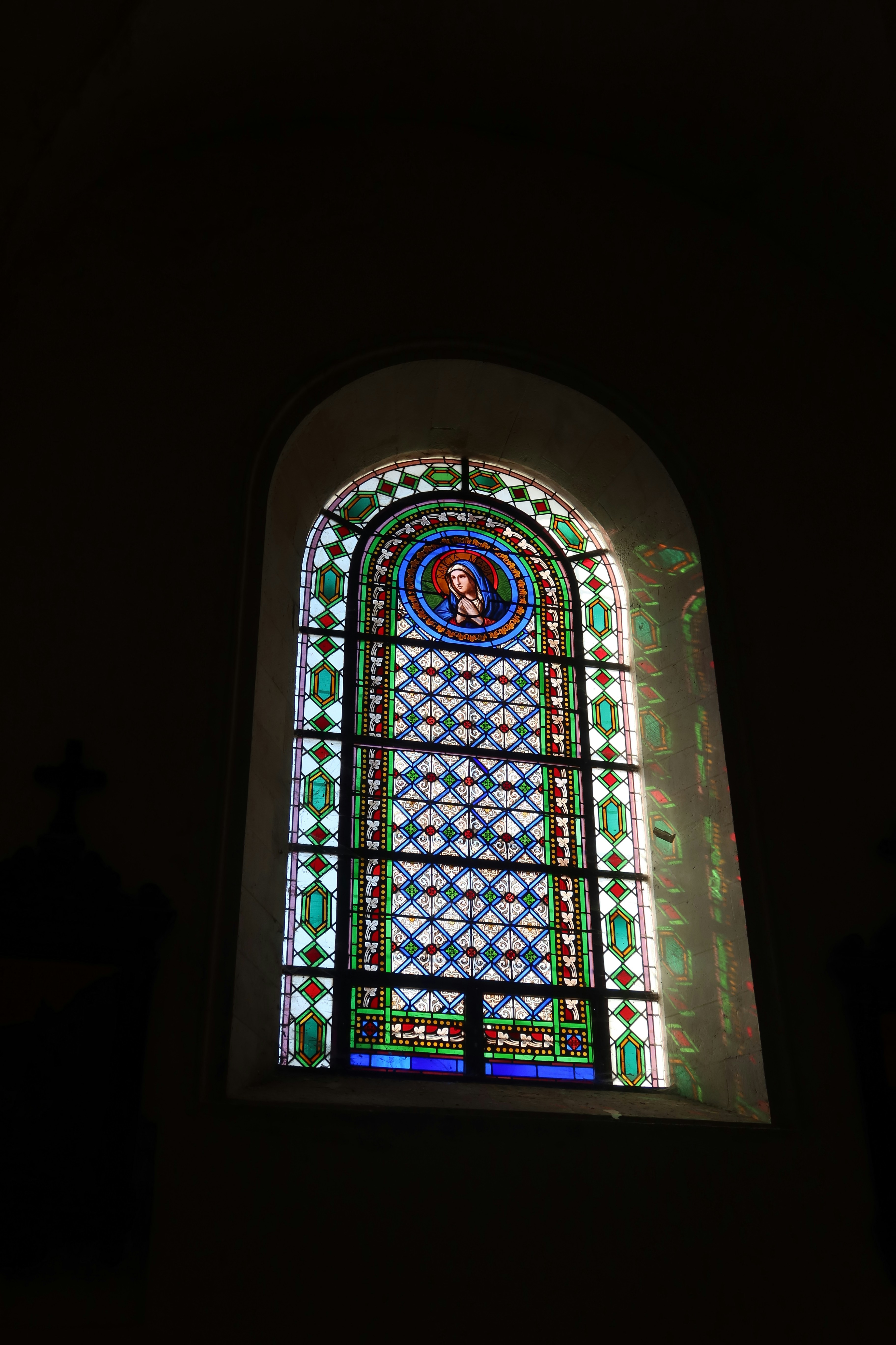 A tall arched stained‑glass window set in a dim church interior, featuring a central medallion depicting the Virgin Mary above an intricate grid of blue, green, and red geometric patterns, with colored light faintly reflecting onto the surrounding stone. | Stained glass window glows in a dark room.
