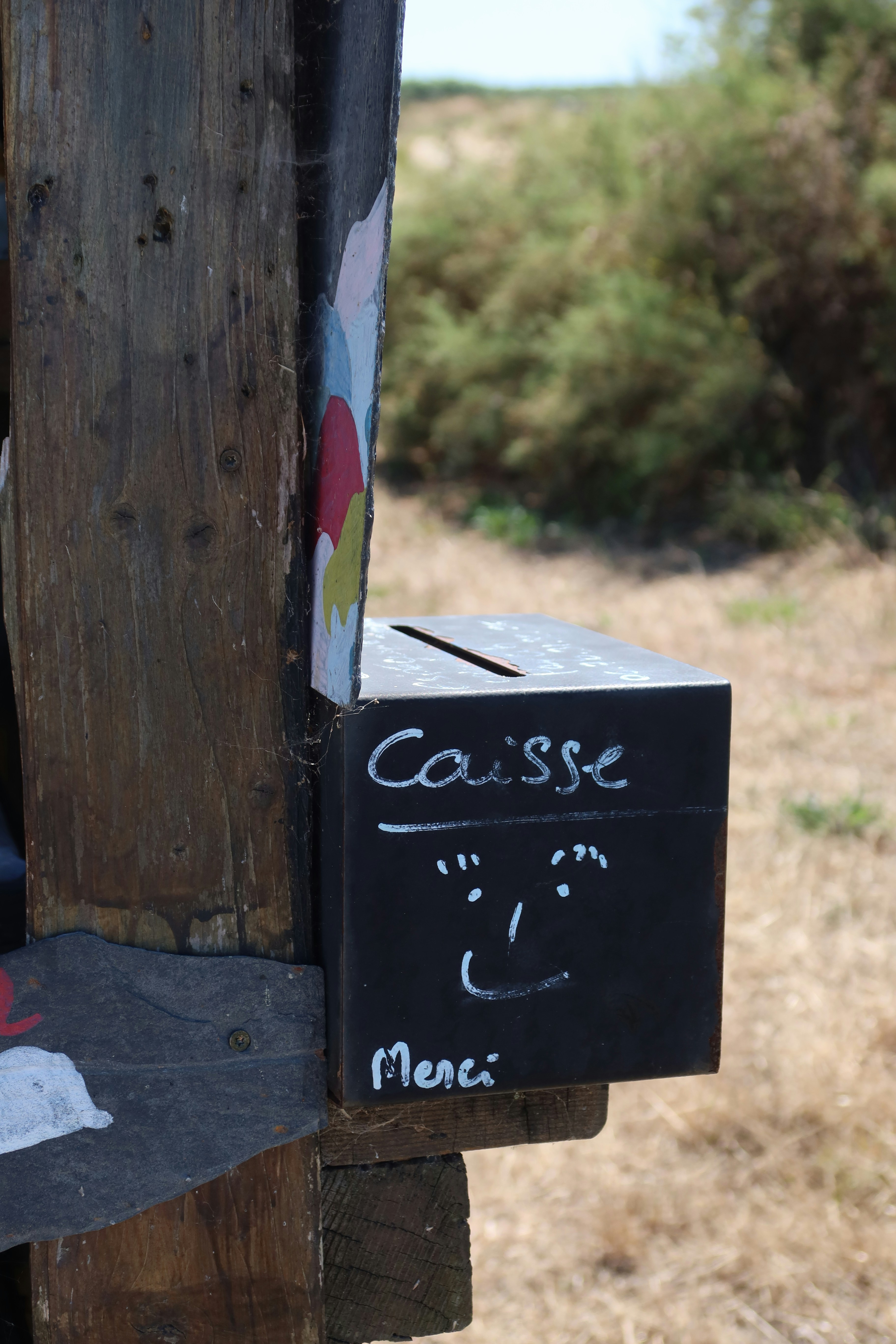 A black metal “Caisse” box with a hand‑drawn smiley face and “Merci” scribbled below is mounted on a weathered wooden post beside sun‑bleached marshland, serving as an honor‑system cash box for a roadside salt stand. | A small donation box with the word "merci".