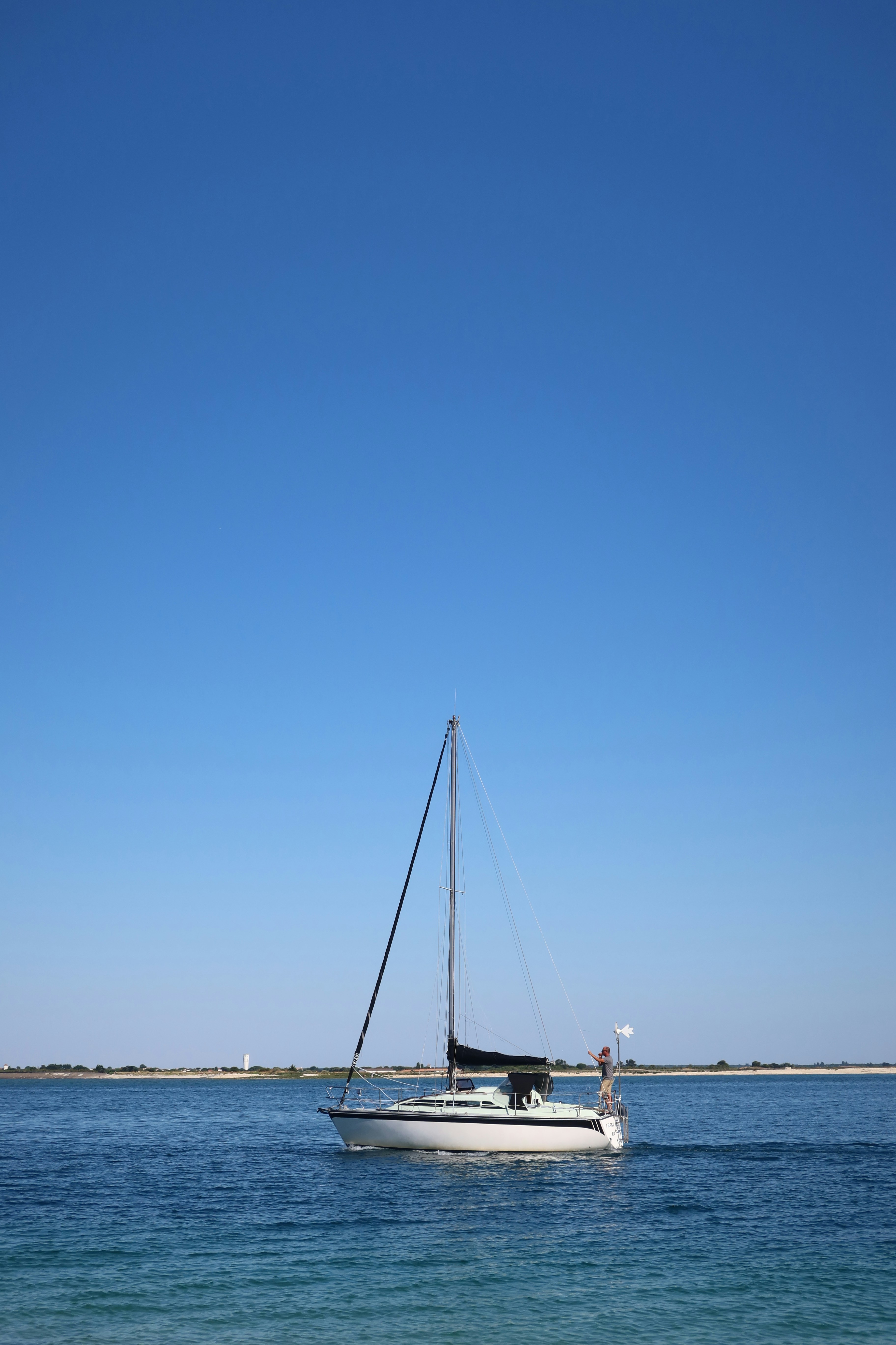 A small white sailboat with a tall mast glides across calm, deep-blue water under a clear sky. A person stands on deck adjusting the rigging, and a low sandy shoreline stretches across the horizon in the distance. | A sailboat sails on a bright, blue sea.