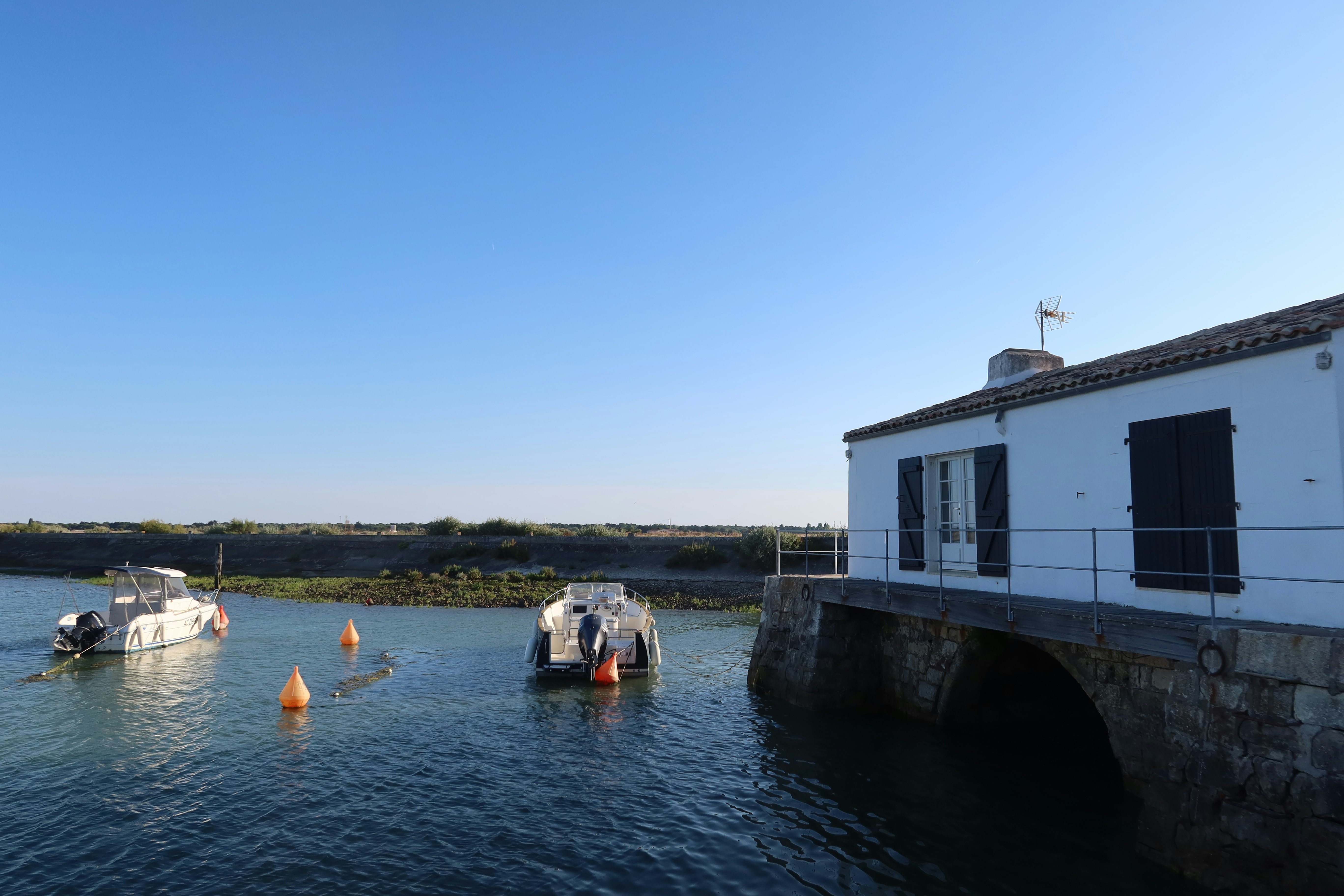 A serene harbor scene at dusk: two small motorboats tied to orange buoys float in the calm, turquoise water alongside a whitewashed boathouse with dark shutters, which sits atop a stone quay featuring an arched opening. Beyond, a low, grassy shoreline stretches under a clear sky transitioning to evening. | Boats are docked at a harbor on a sunny day.
