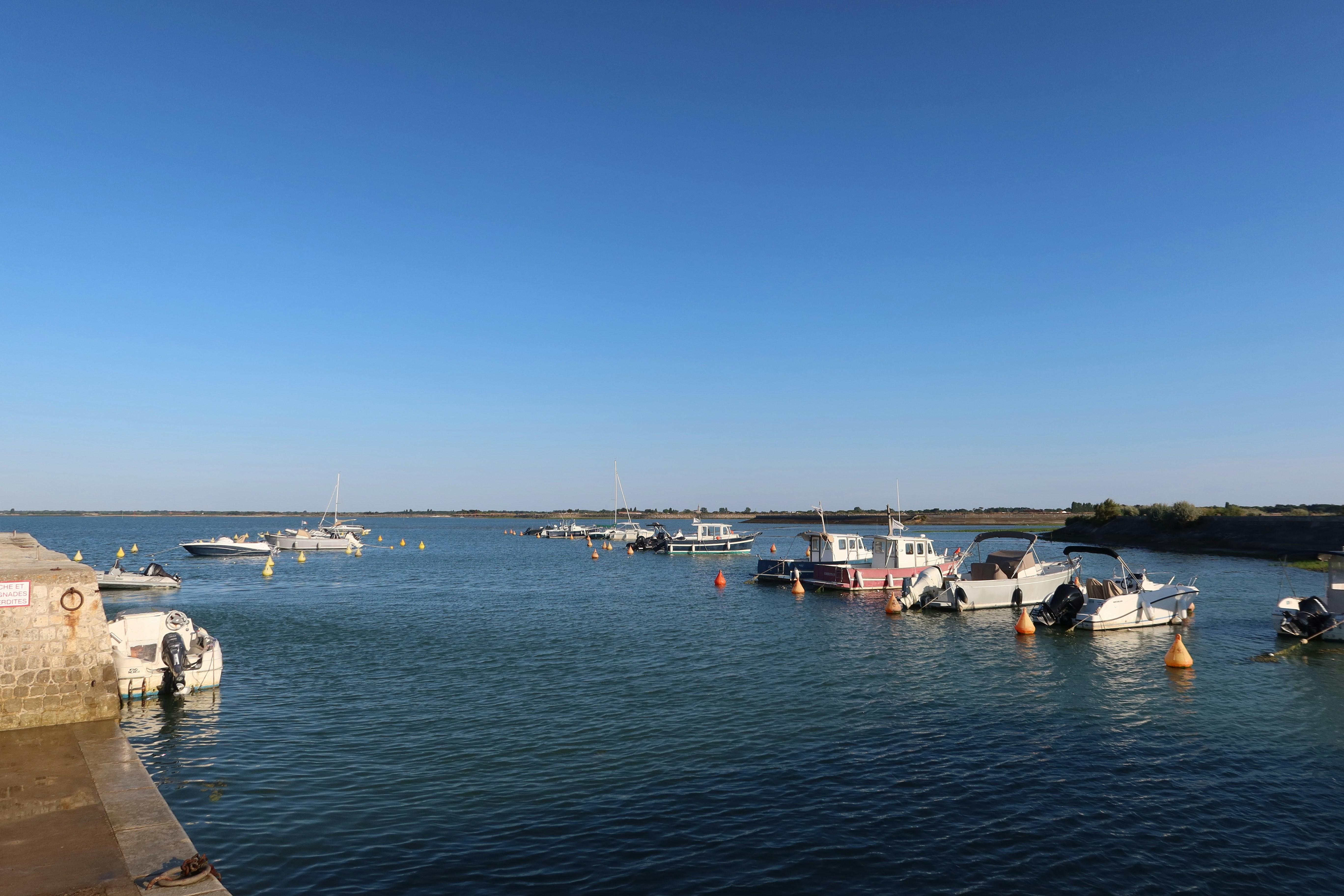 A tranquil seaside harbor at dusk, with small motorboats moored to bright orange buoys beside a stone quay and waterfront building, set against calm turquoise water and a clear evening sky. | Boats are moored in a calm, sunny harbor.