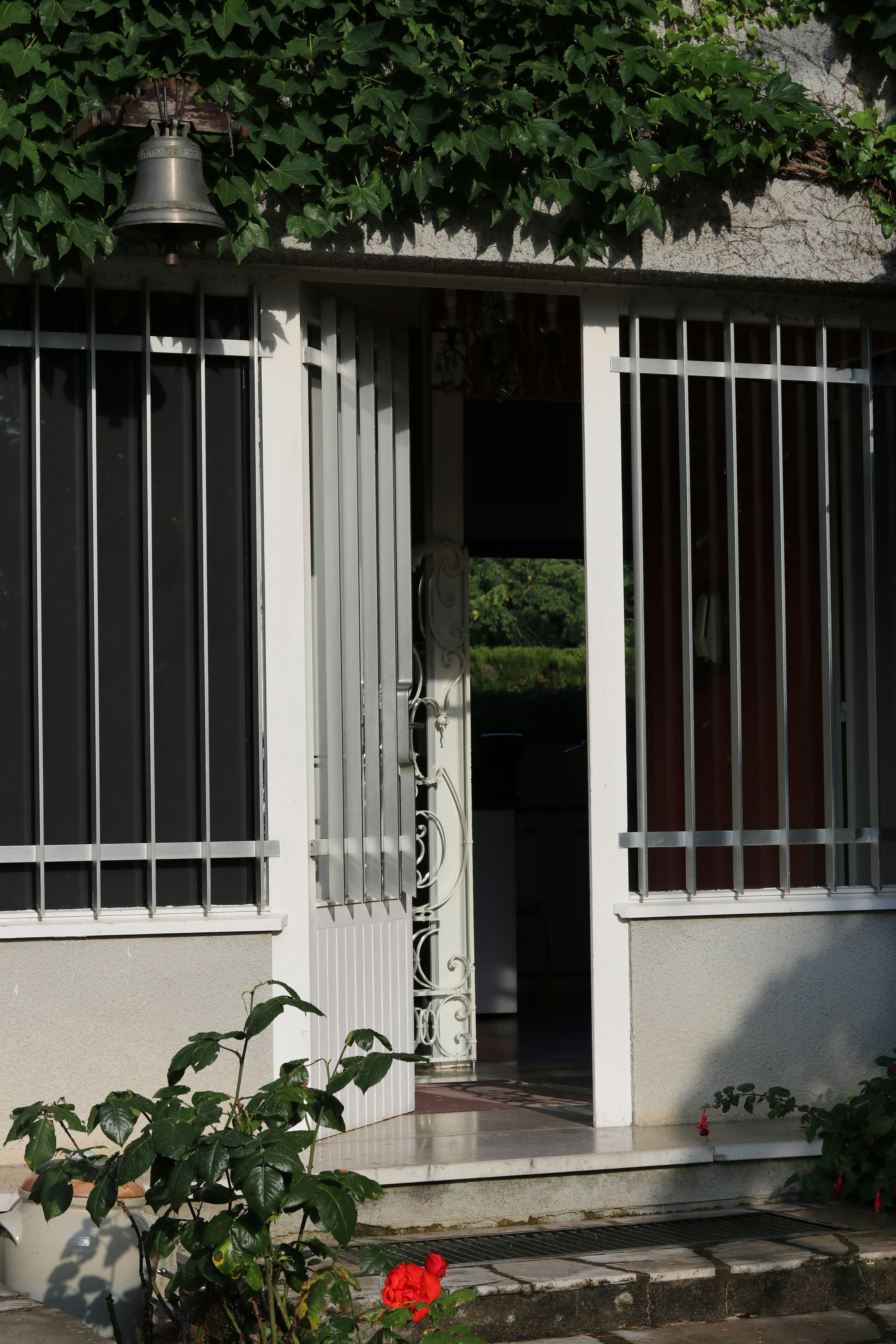 A white metal‑barred doorway opens beneath a canopy of climbing ivy and an antique brass bell, with a vibrant red rose in a pot at the threshold, inviting a glimpse into a sunlit home interior. | Open doorway with bars, bell, and climbing vines.
