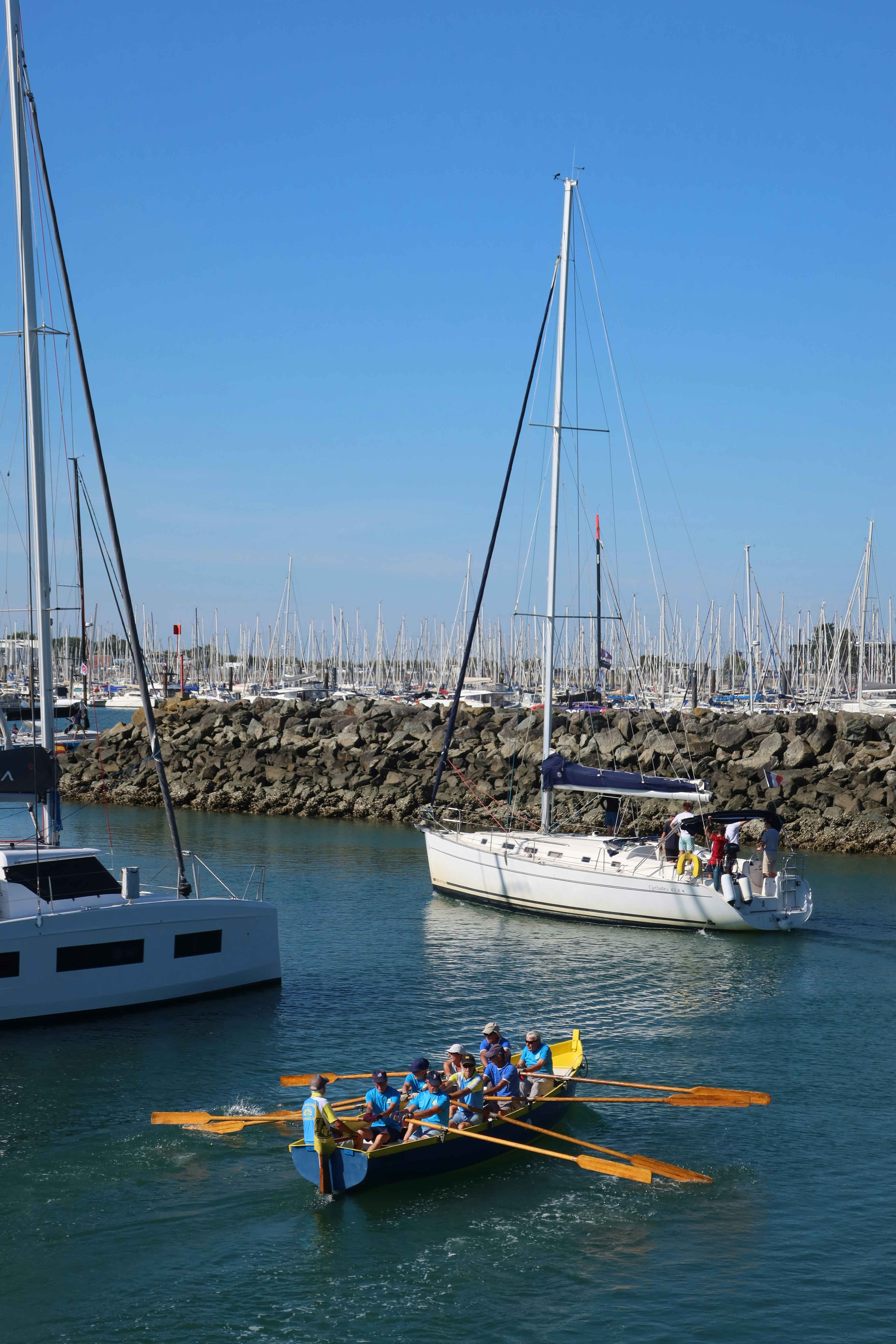 A traditional long rowing boat with a team of eight rowers in blue shirts and yellow life vests, each handling a wooden oar, gliding through a calm marina channel lined by white sailboats, a rocky breakwater, and clear blue sky. | Rowing crew and sailboats on a sunny day.