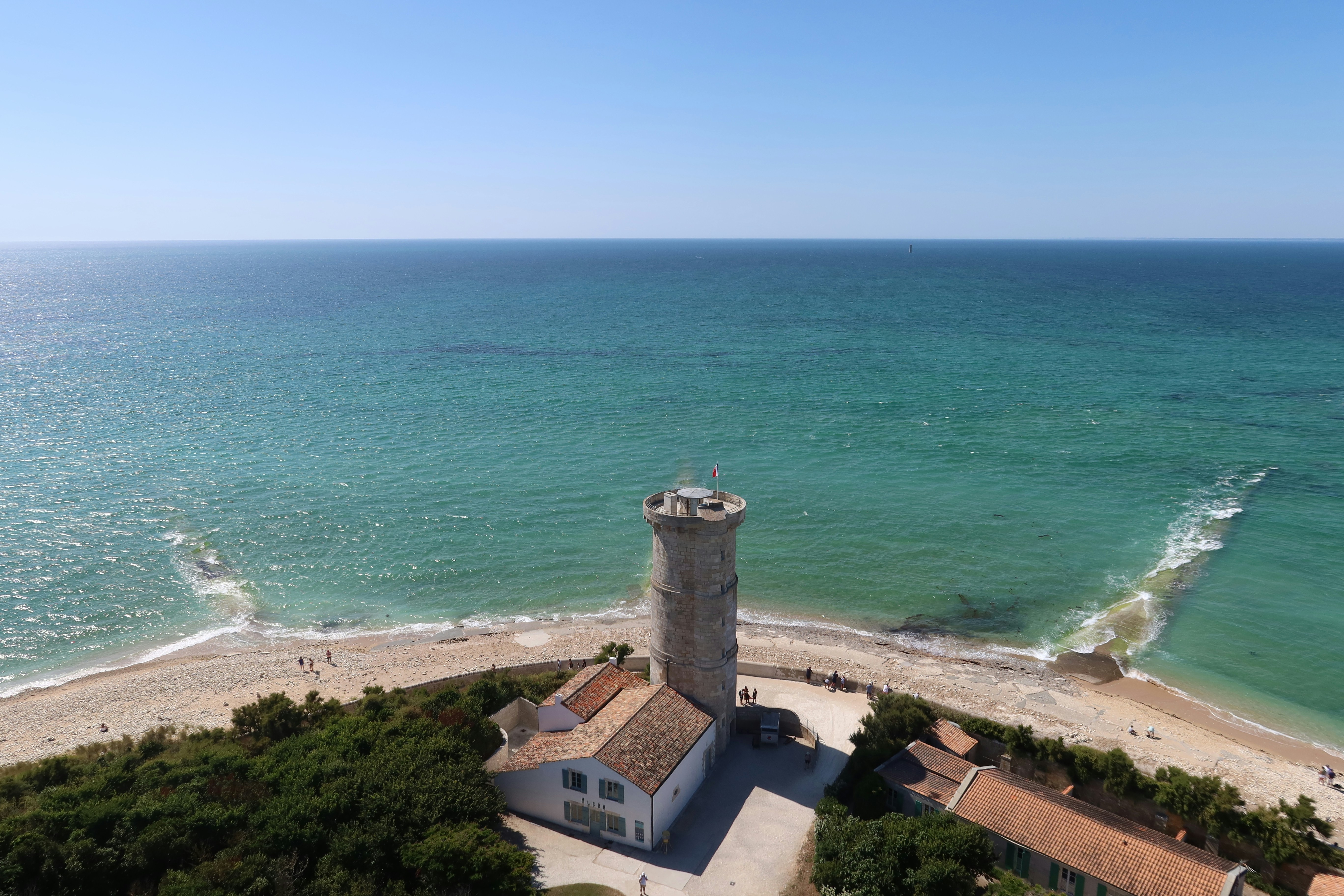 A tall stone lighthouse tower beside red‑tiled buildings on a sandy beach, overlooking turquoise Atlantic waters under a clear blue sky. | A lighthouse stands guard over the sea.