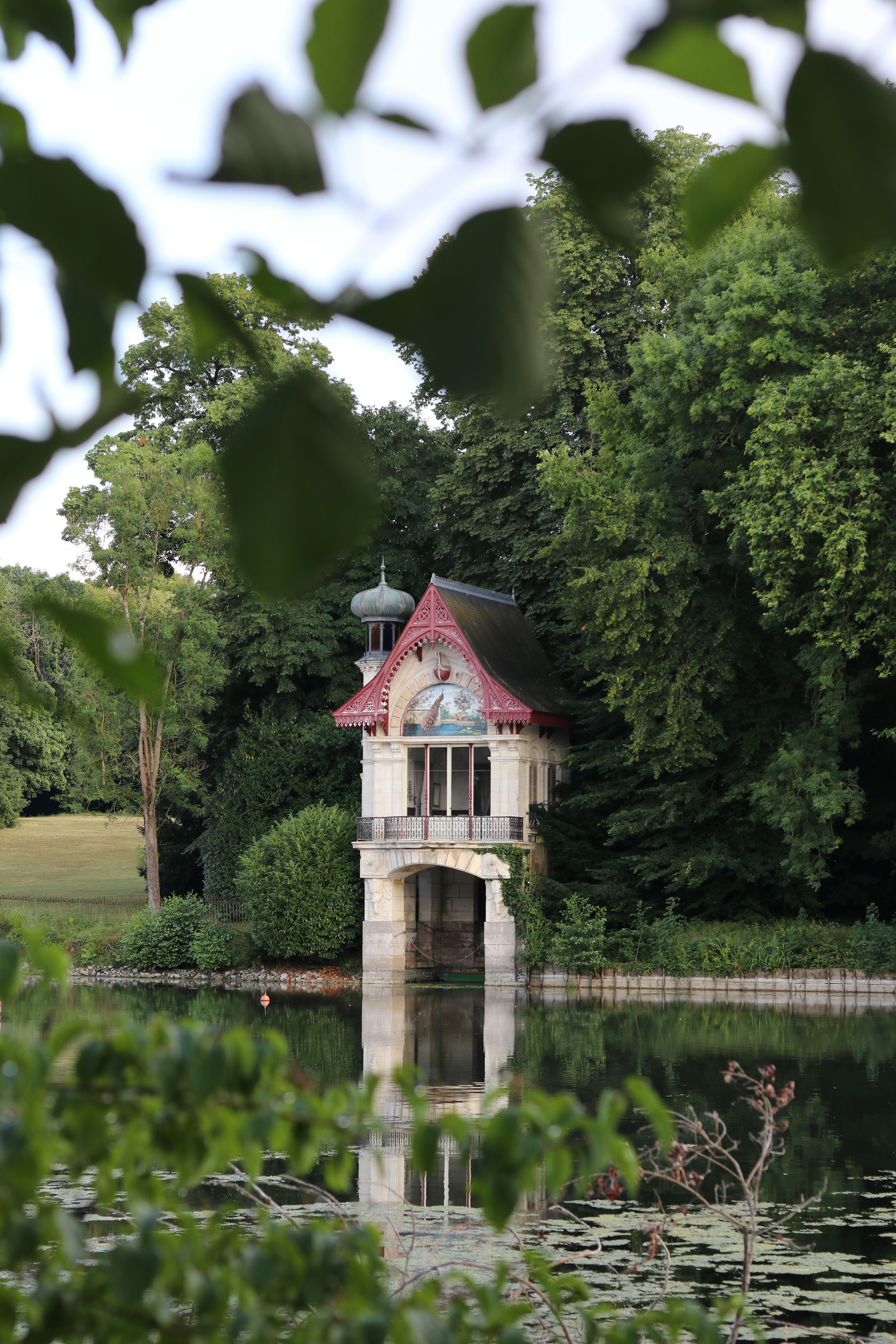 A romantic boathouse with a decorative red gabled roof and painted mosaic panel stands on stone pillars at the edge of a tranquil lake, its ornate facade and turret reflected in the water and framed by lush green foliage. | A charming boathouse reflects in a calm lake.