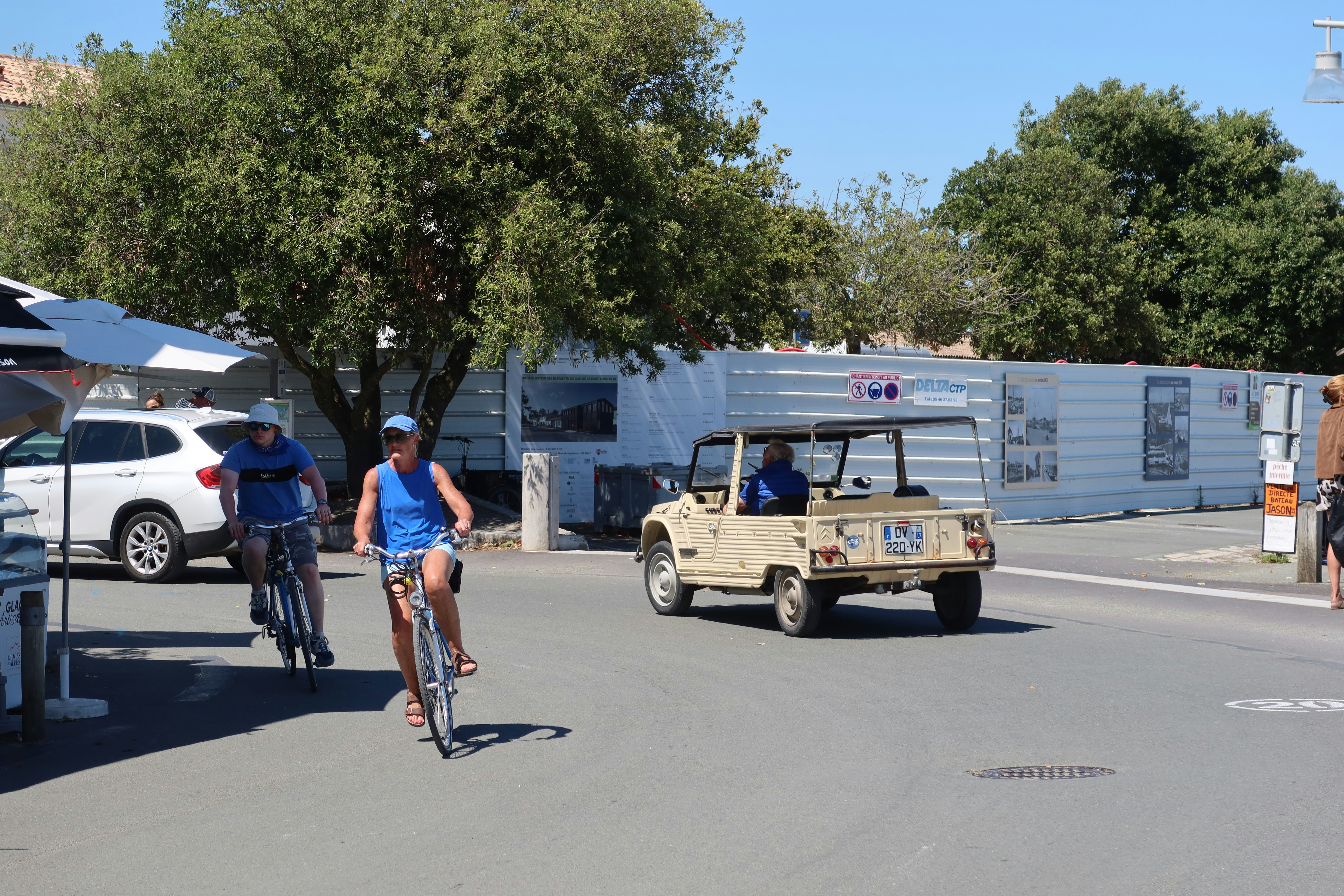 Two cyclists in casual summer attire ride past a beige Citroën Méhari cruising down a sunlit harbor street, flanked by parked cars, leafy trees, and a construction fence under a clear blue sky. | People are biking and driving on a sunny day.
