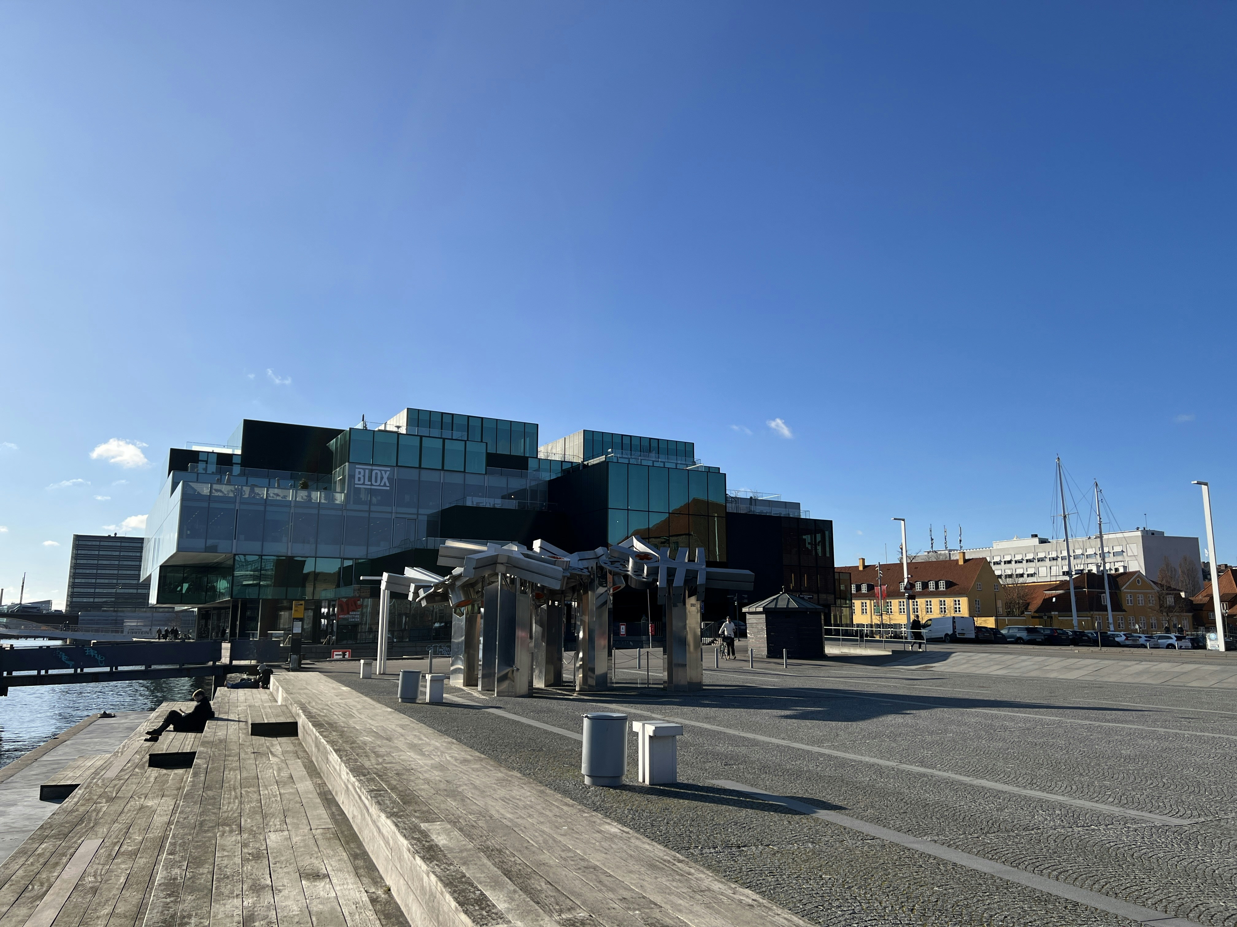 Grand urban structure with expansive glass facade dominating the Copenhagen waterfront. | Modern building stands on a waterfront with a blue sky.