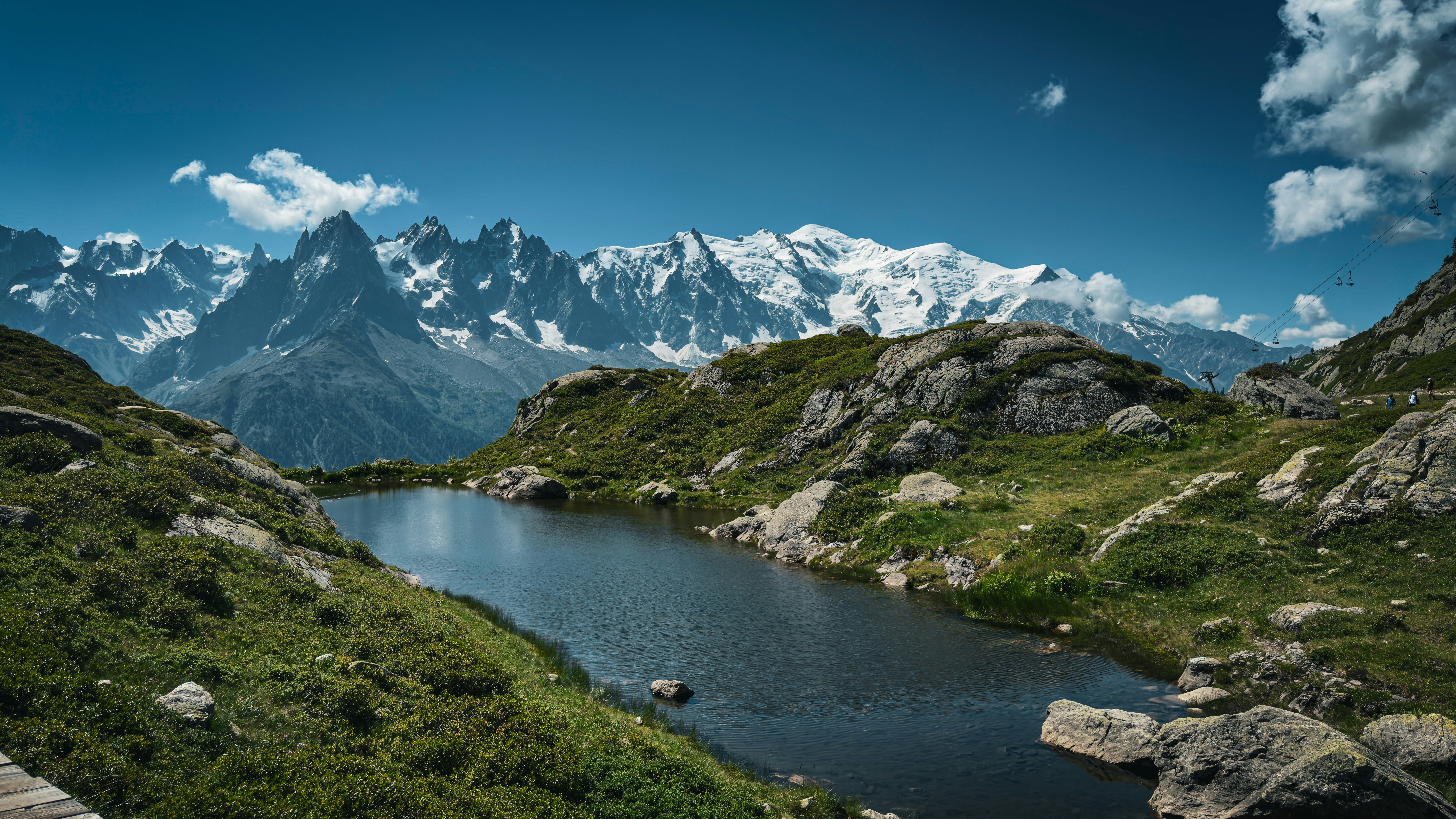 Lush green hills frame a tranquil pond, reflecting the towering snow-capped mountains under a clear blue sky.