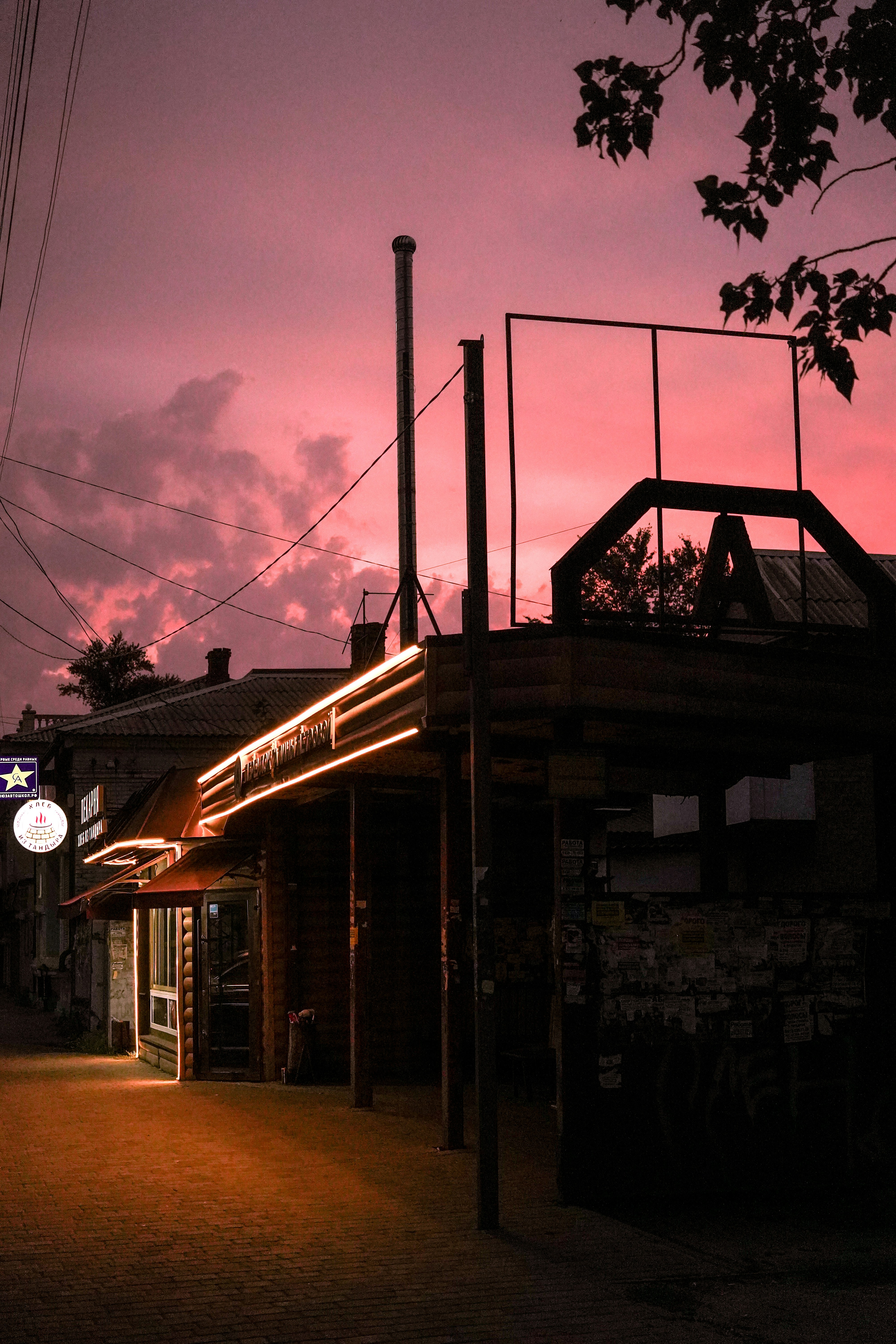 A cozy street scene illuminated by warm lights against a vibrant pink twilight sky, showcasing a rustic building with signage and posters.