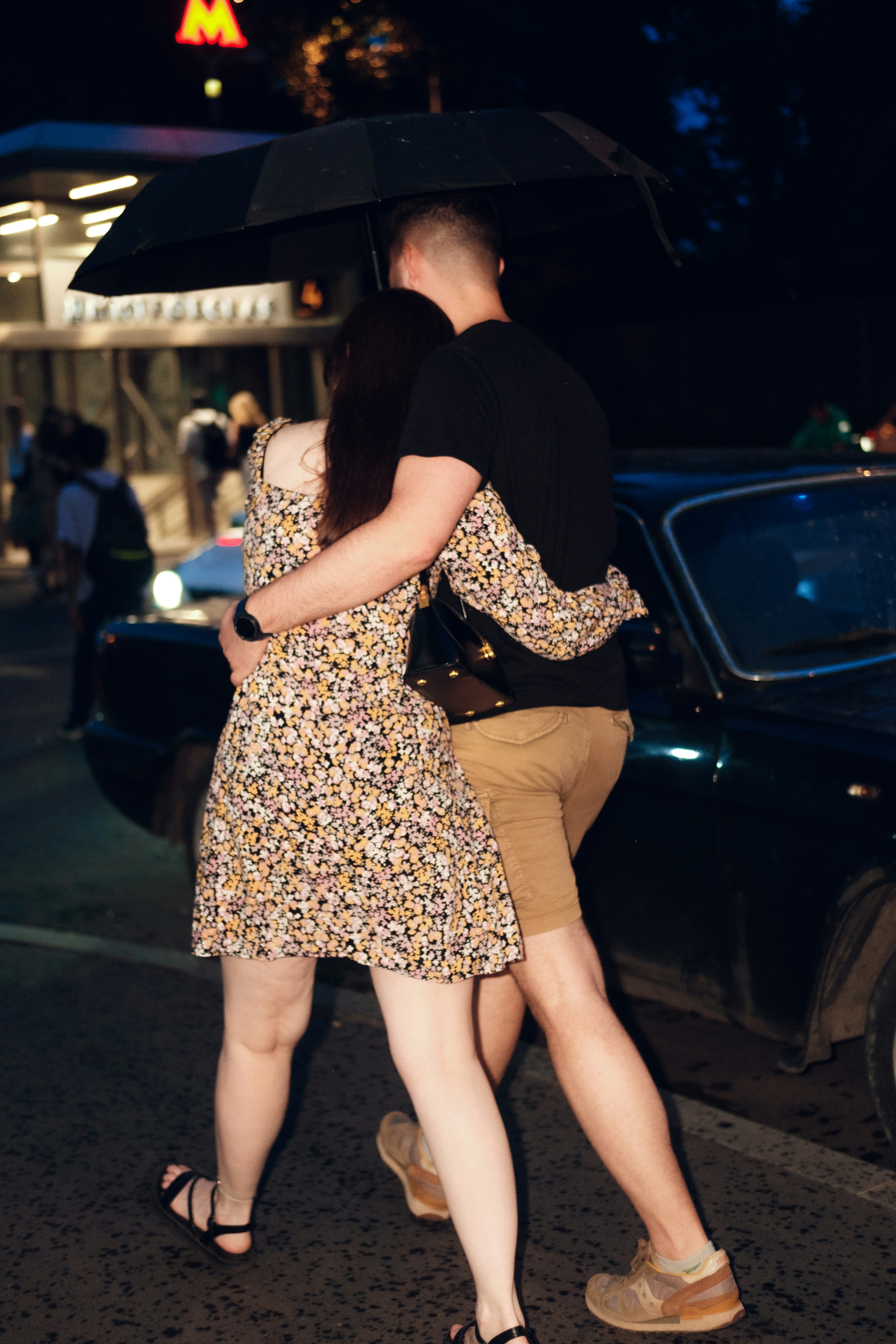 Couple sharing an umbrella while walking on a city street at night, illuminated by soft streetlights and a nearby sign.