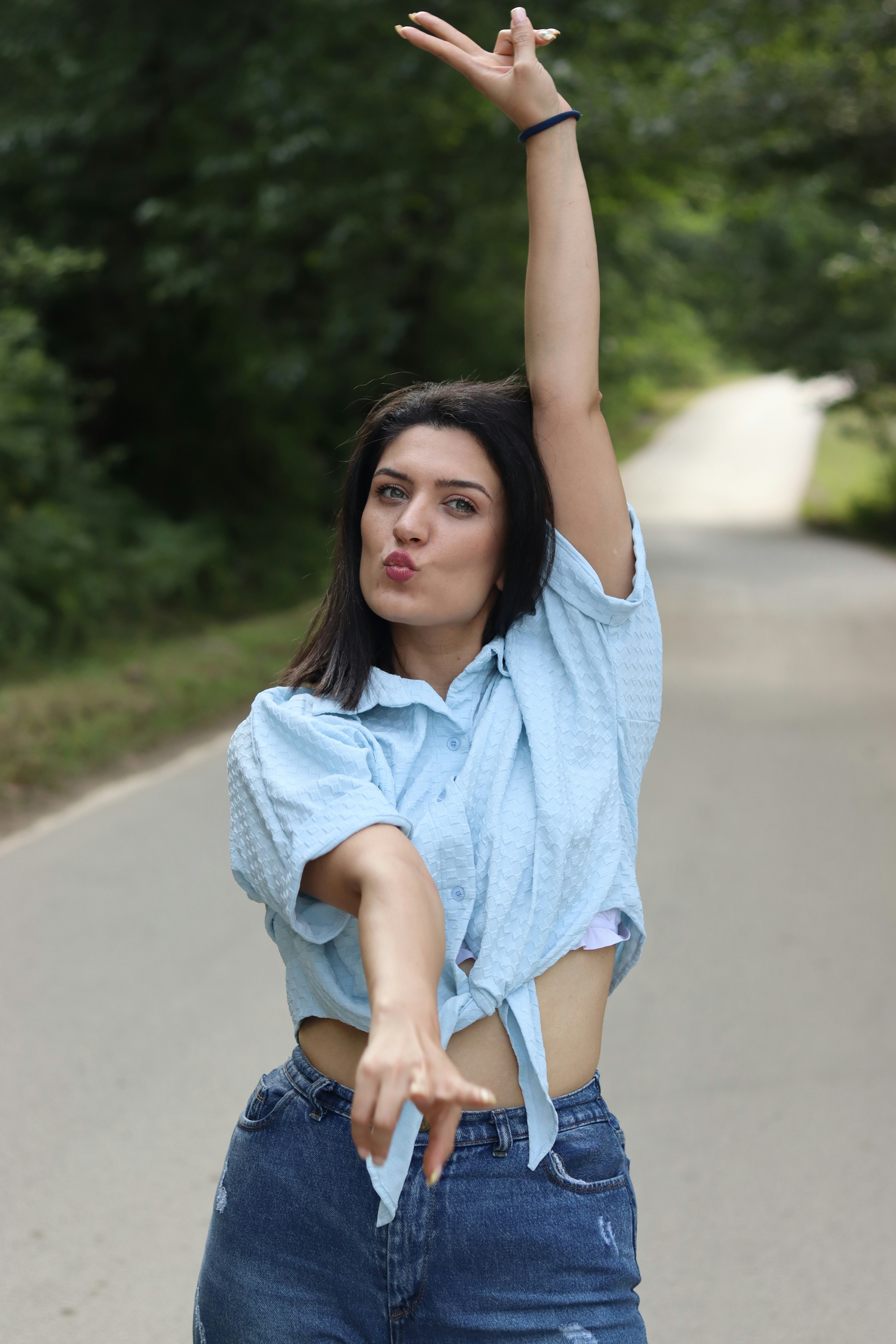 Young woman playfully poses with a raised arm on a serene country road, surrounded by greenery.