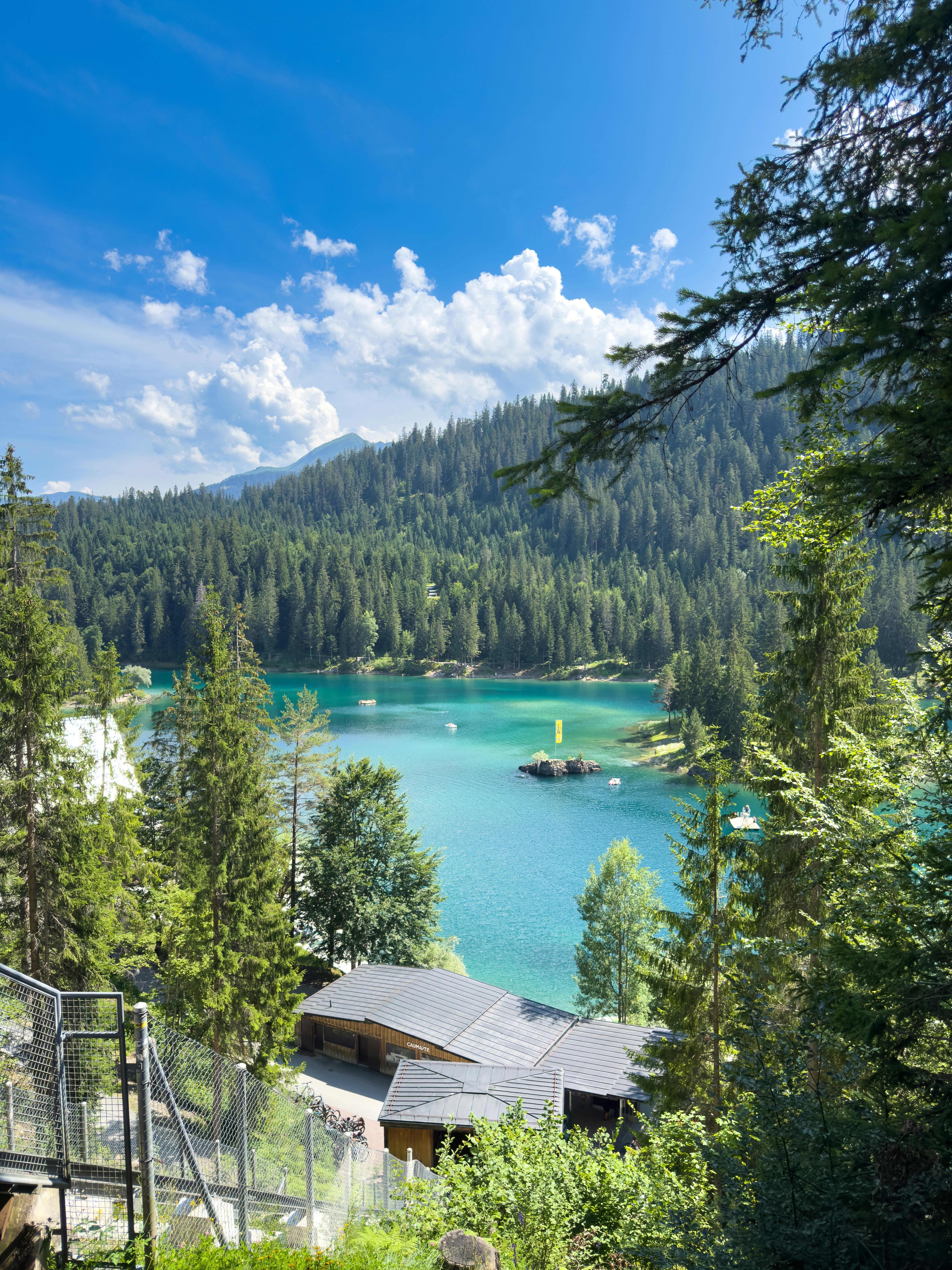 A serene alpine lake surrounded by towering pine trees, with a vibrant turquoise hue and distant mountains under a partly cloudy sky.