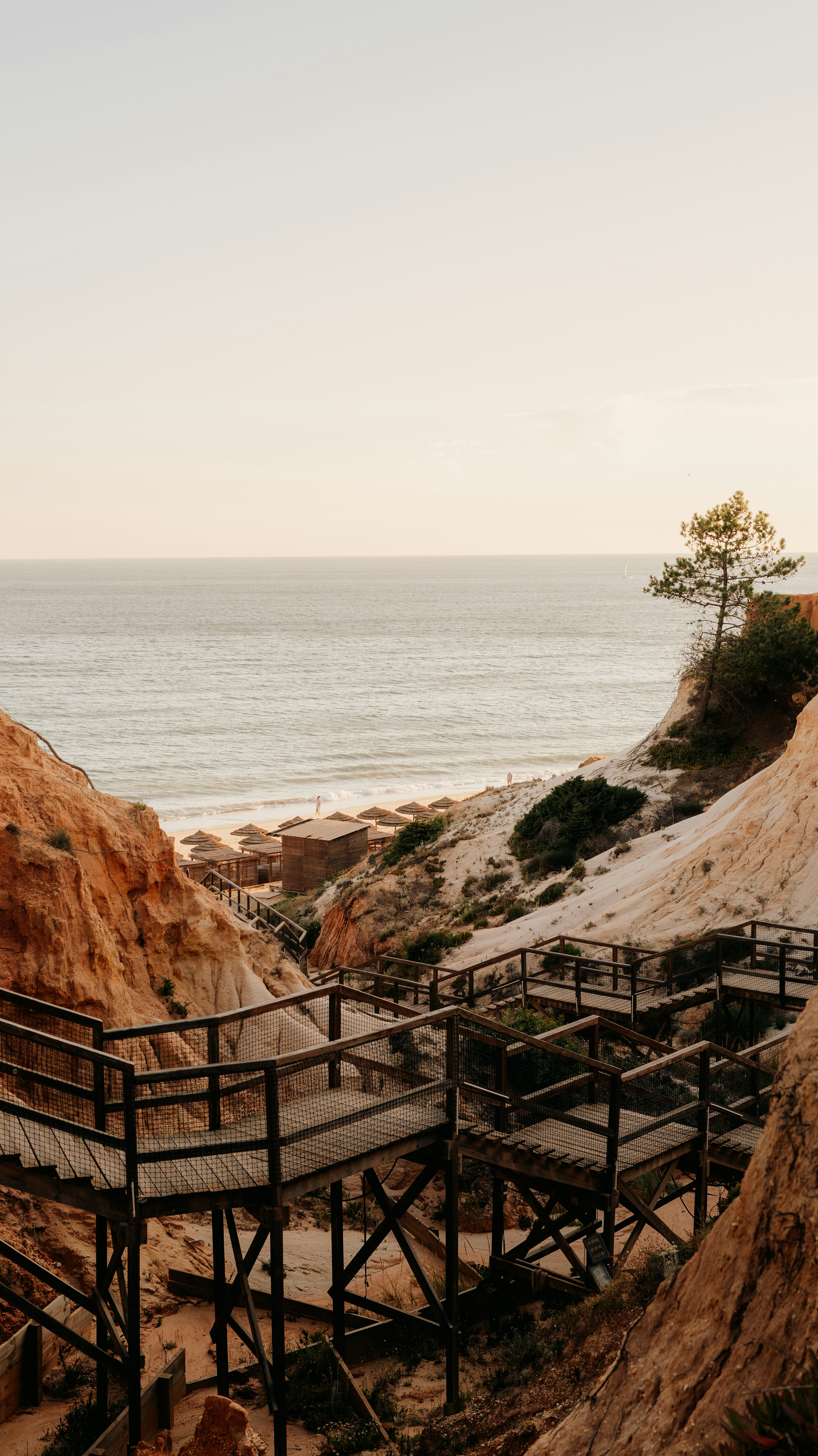 Wooden walkway meanders through rugged cliffs leading to a tranquil beach at sunset.