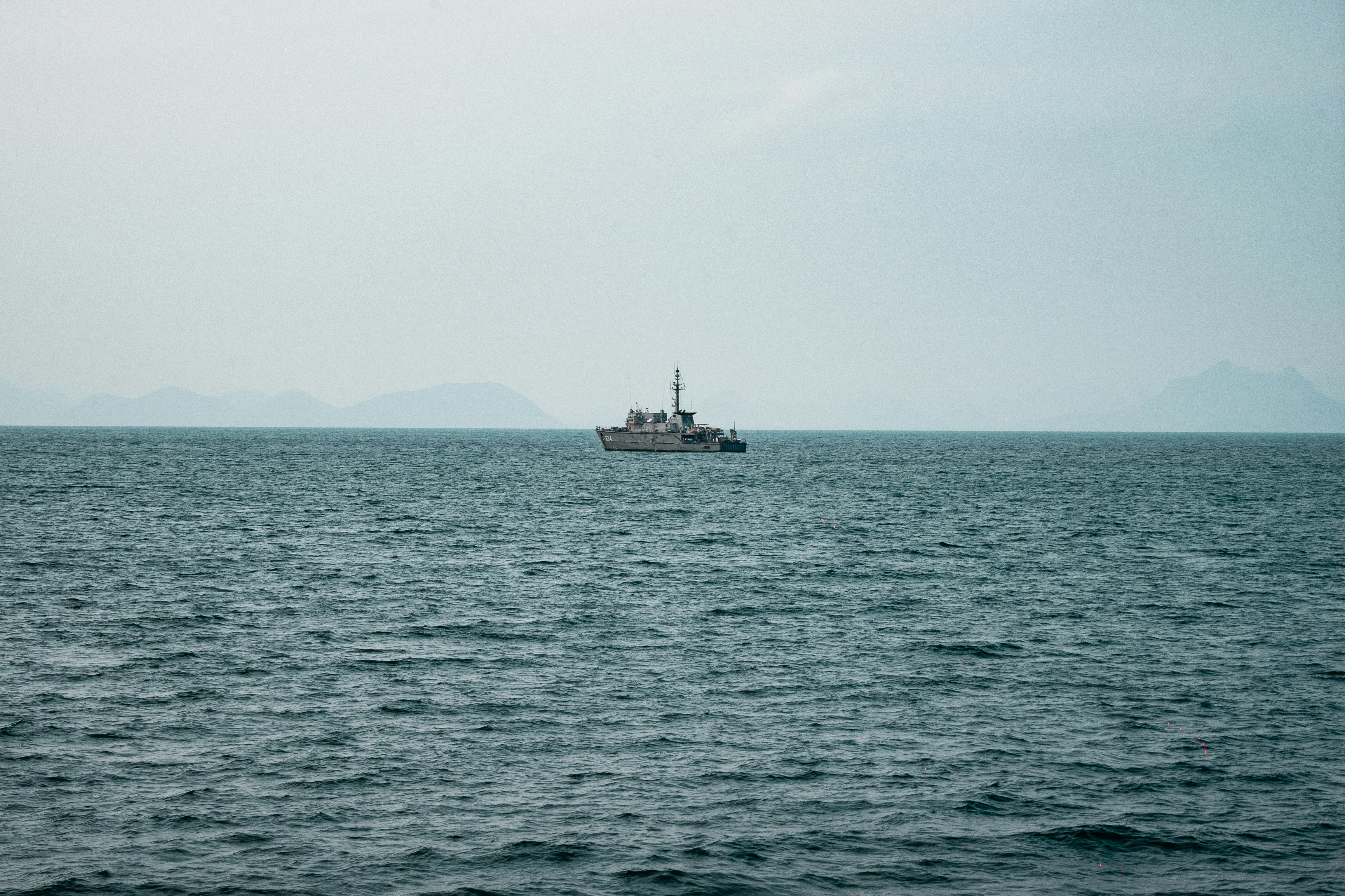 A lone ship navigating the calm sea, framed by distant mountains under a soft, overcast sky.