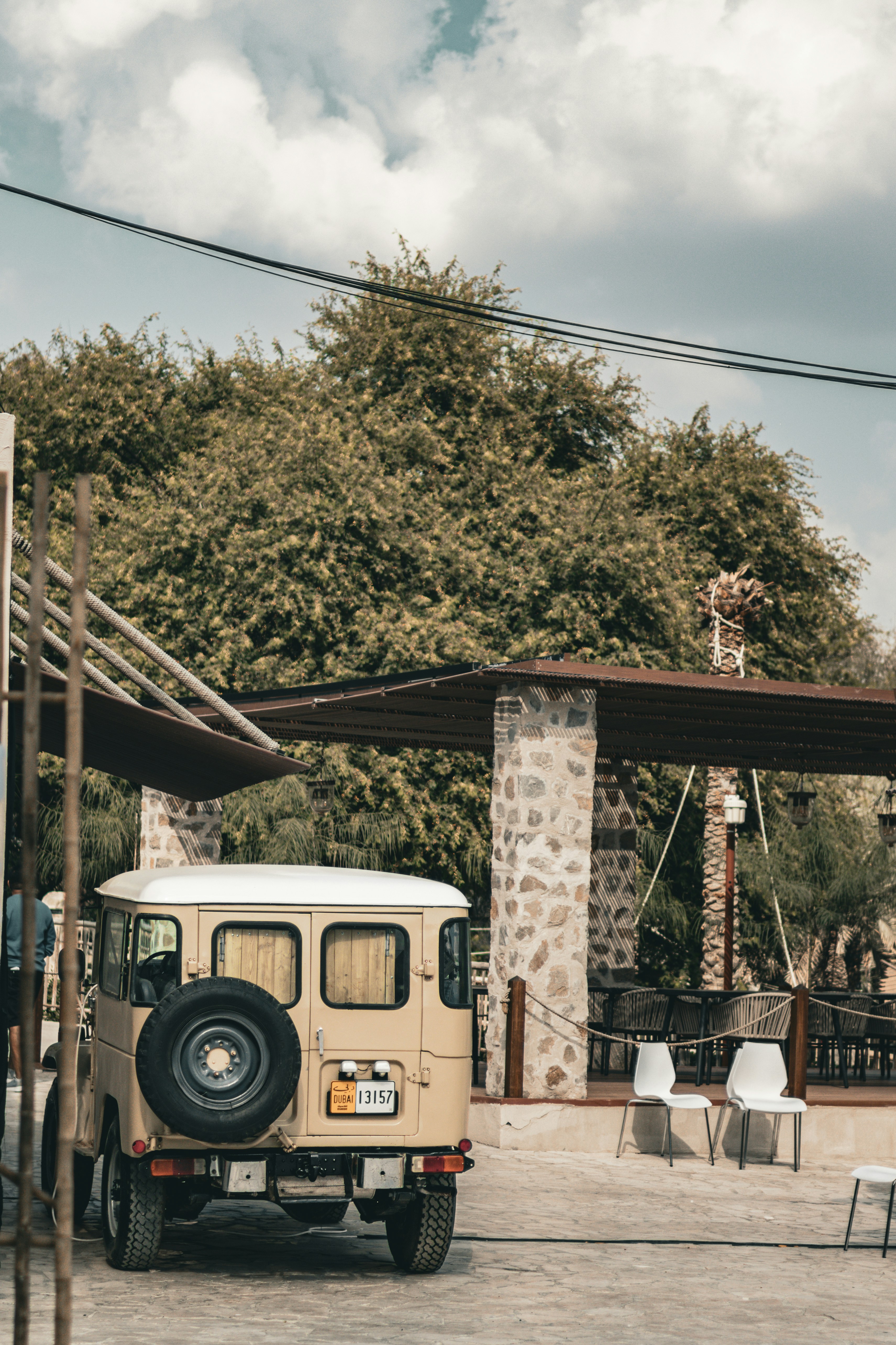 A vintage beige off-road vehicle, possibly a Toyota Land Cruiser, is parked in a rustic courtyard adorned with stone pillars and shaded roofing. The Dubai license plate adds regional character, while the composition exudes a nostalgic, explorer vibe. Shot in natural daylight with a desaturated, earthy tone palette, the image captures a peaceful blend of nature and heritage design. | A vintage jeep parked outside a shaded outdoor seating area.