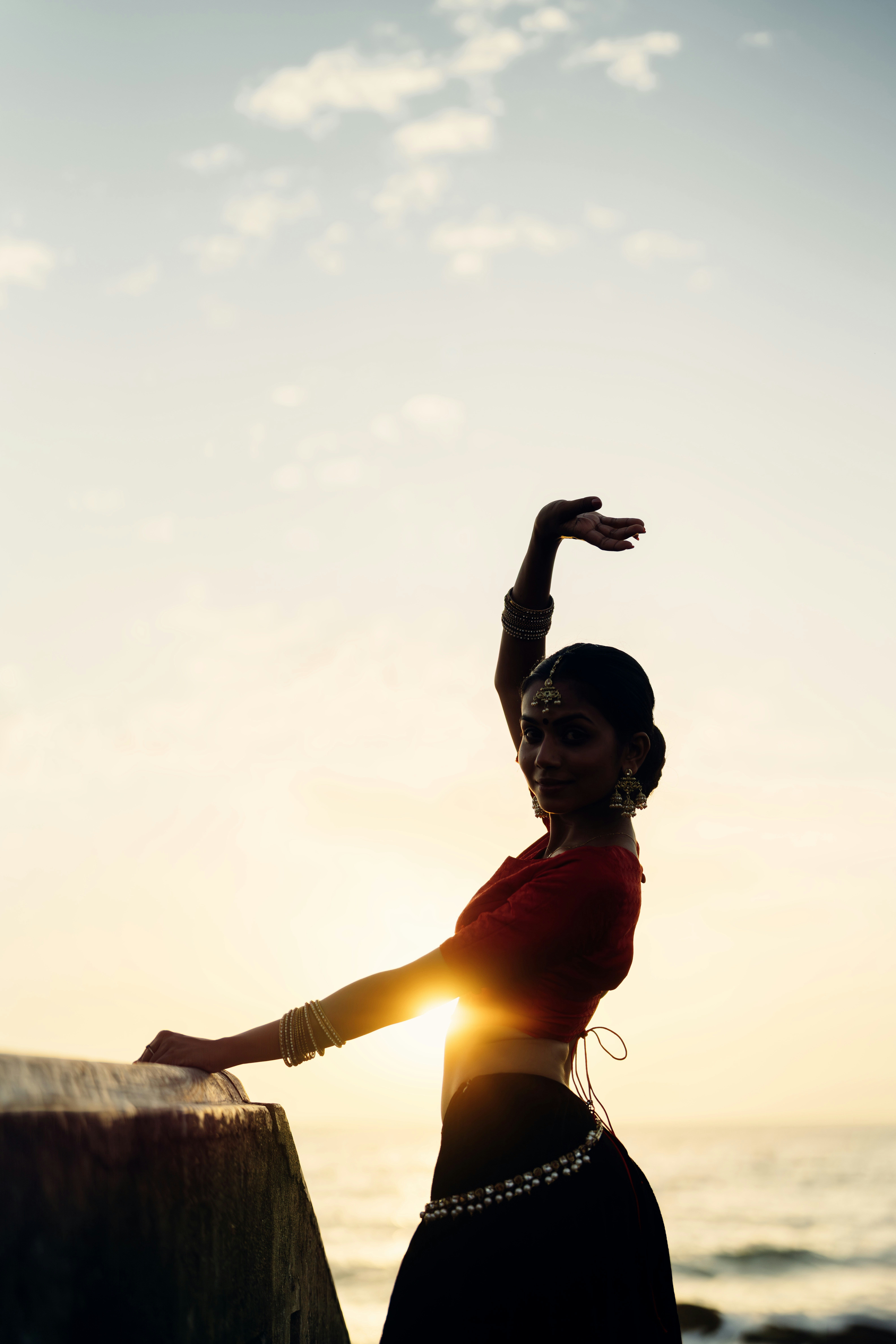 Dancer striking an elegant pose at sunset, showcasing intricate jewelry and traditional attire. The silhouette contrasts beautifully with the glowing sky.