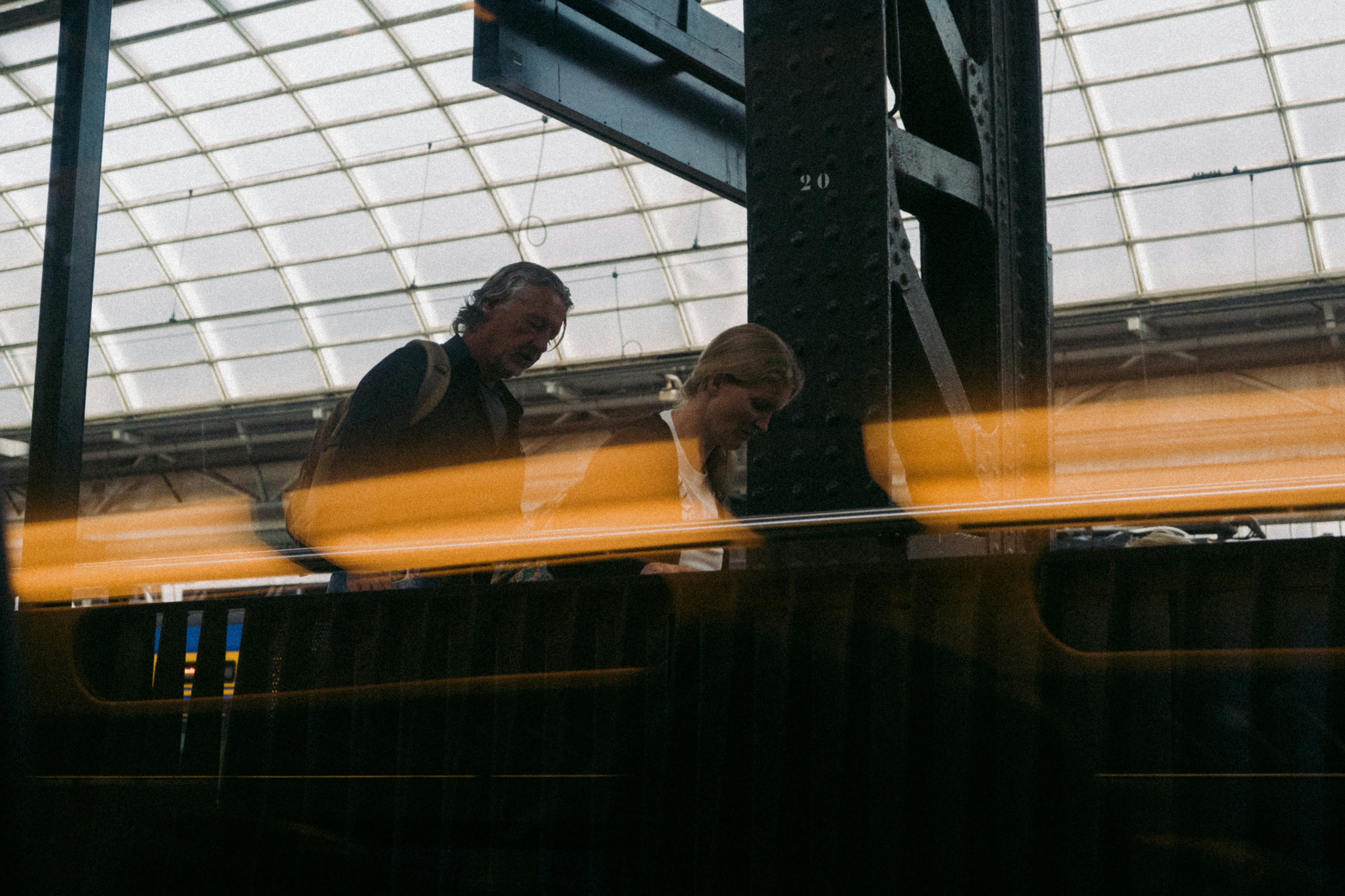 Two commuters absorbed in their devices at a train station, with a blurred train passing by. The scene captures the essence of urban transit.