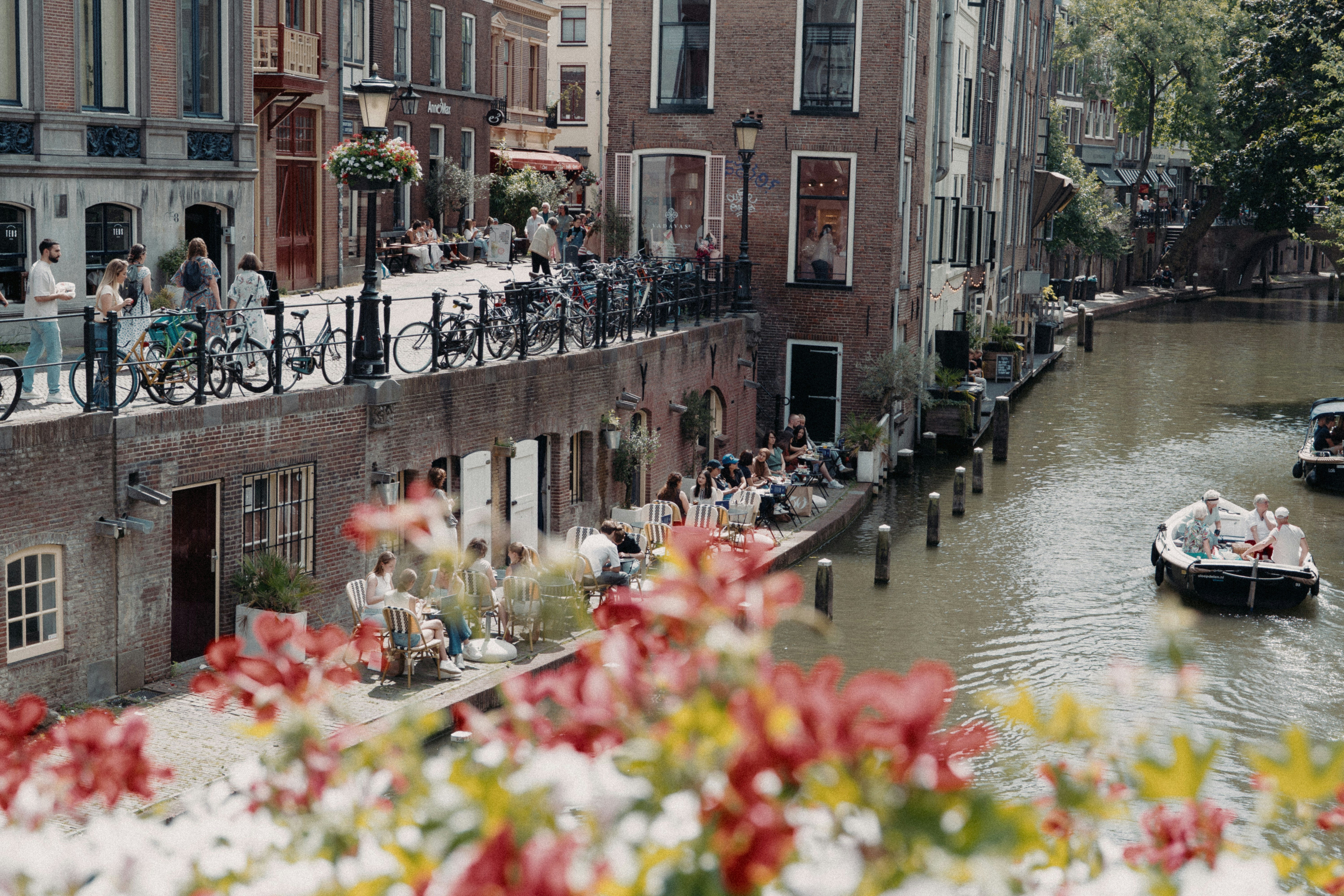 Bustling canal-side terraces spill onto historic quays, with people dining and boats drifting by, all bathed in warm light and framed by vibrant summer flowers—a lively portrait of old-world charm and carefree leisure | A scenic canal view with people enjoying the day.
