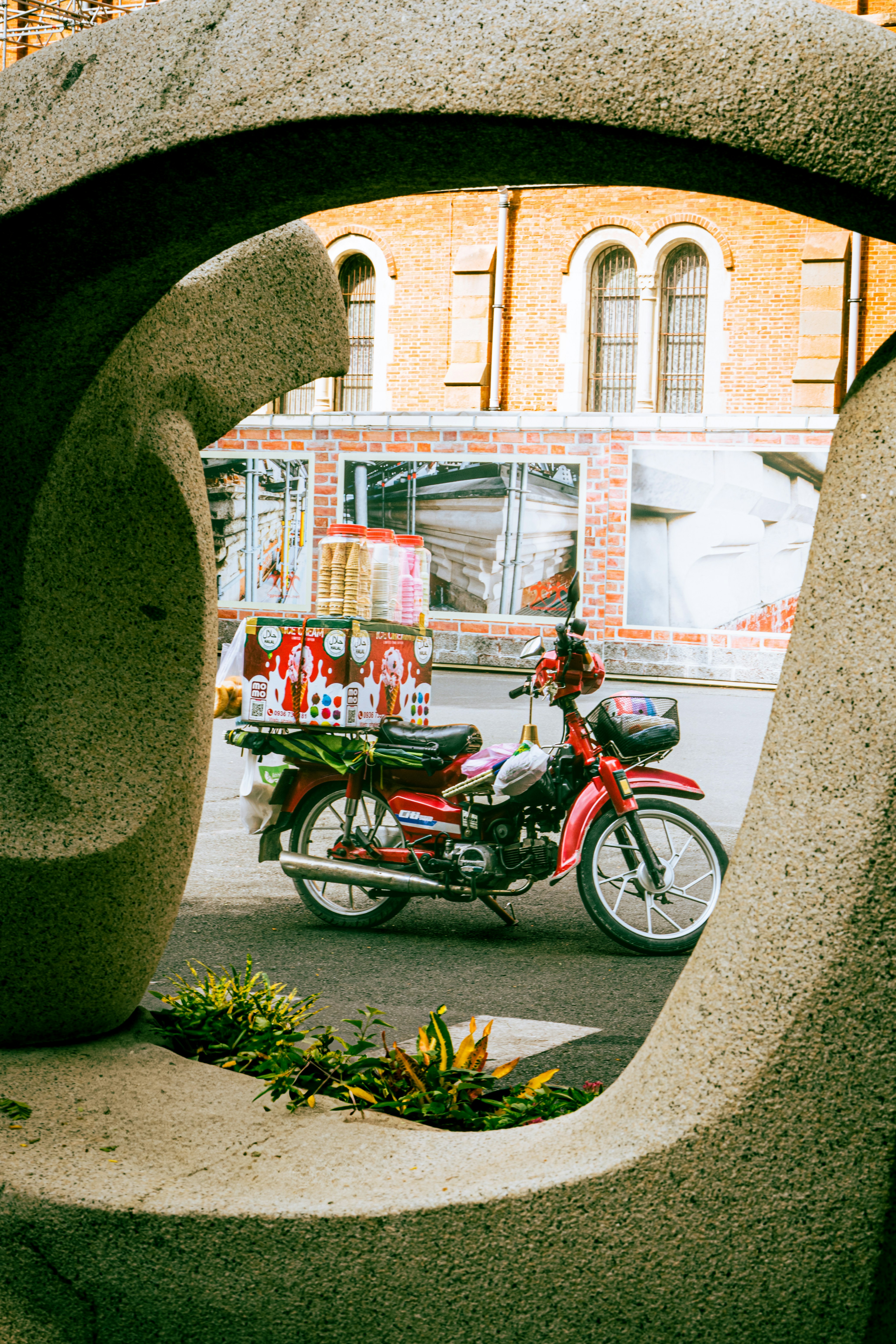 A vibrant red motorcycle laden with colorful goods parked beneath a sculptural arch in a bustling cityscape.