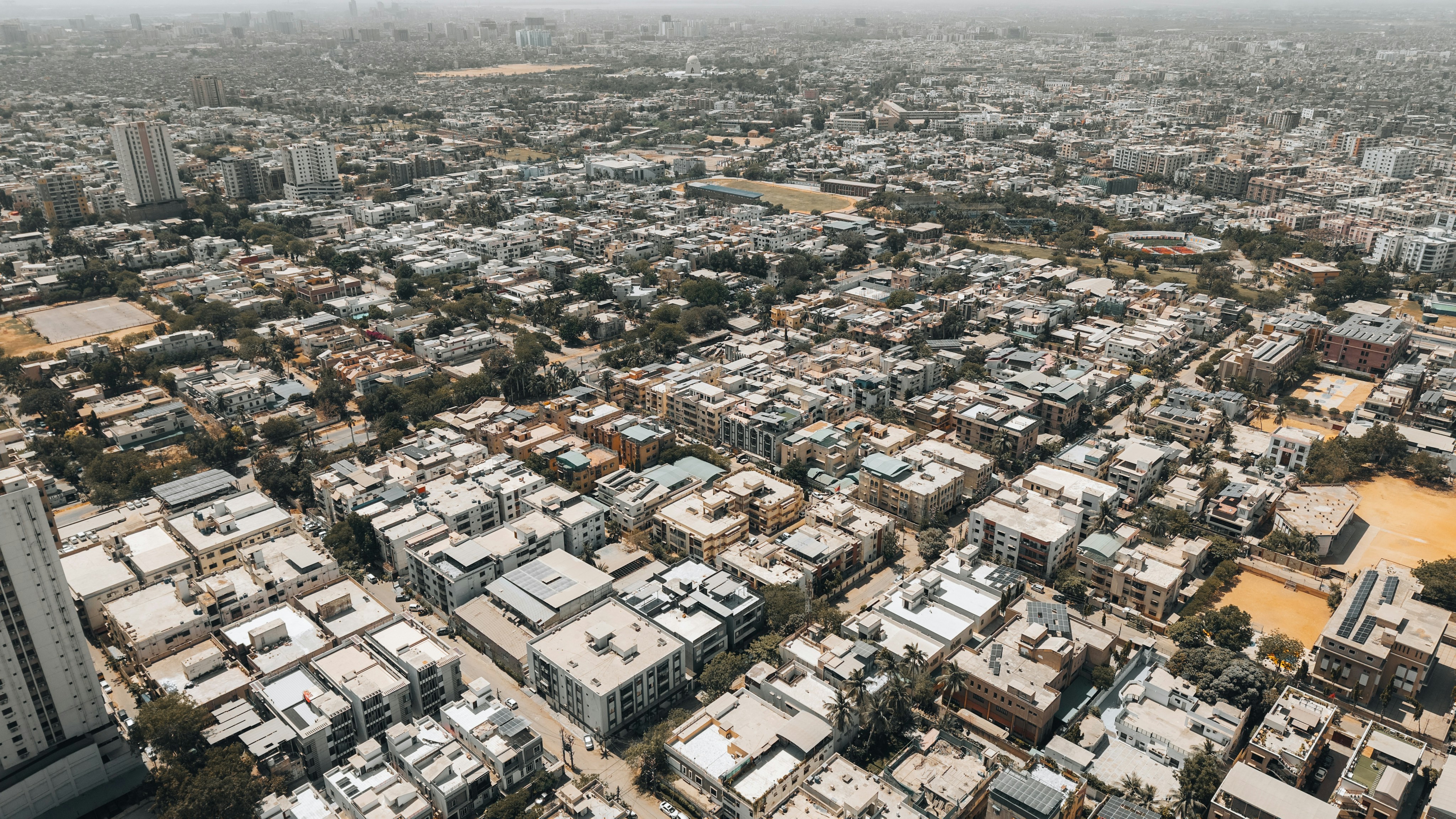 A wide-angle drone photograph capturing the residential blocks and street grid of Karachi, Pakistan. The white dome of Mazar-e-Quaid sits quietly on the skyline, offering a symbolic connection between history and the city’s dense modern sprawl. Edited with a clean cinematic tone for a documentary-style aesthetic. | Aerial view showcases a densely populated city.