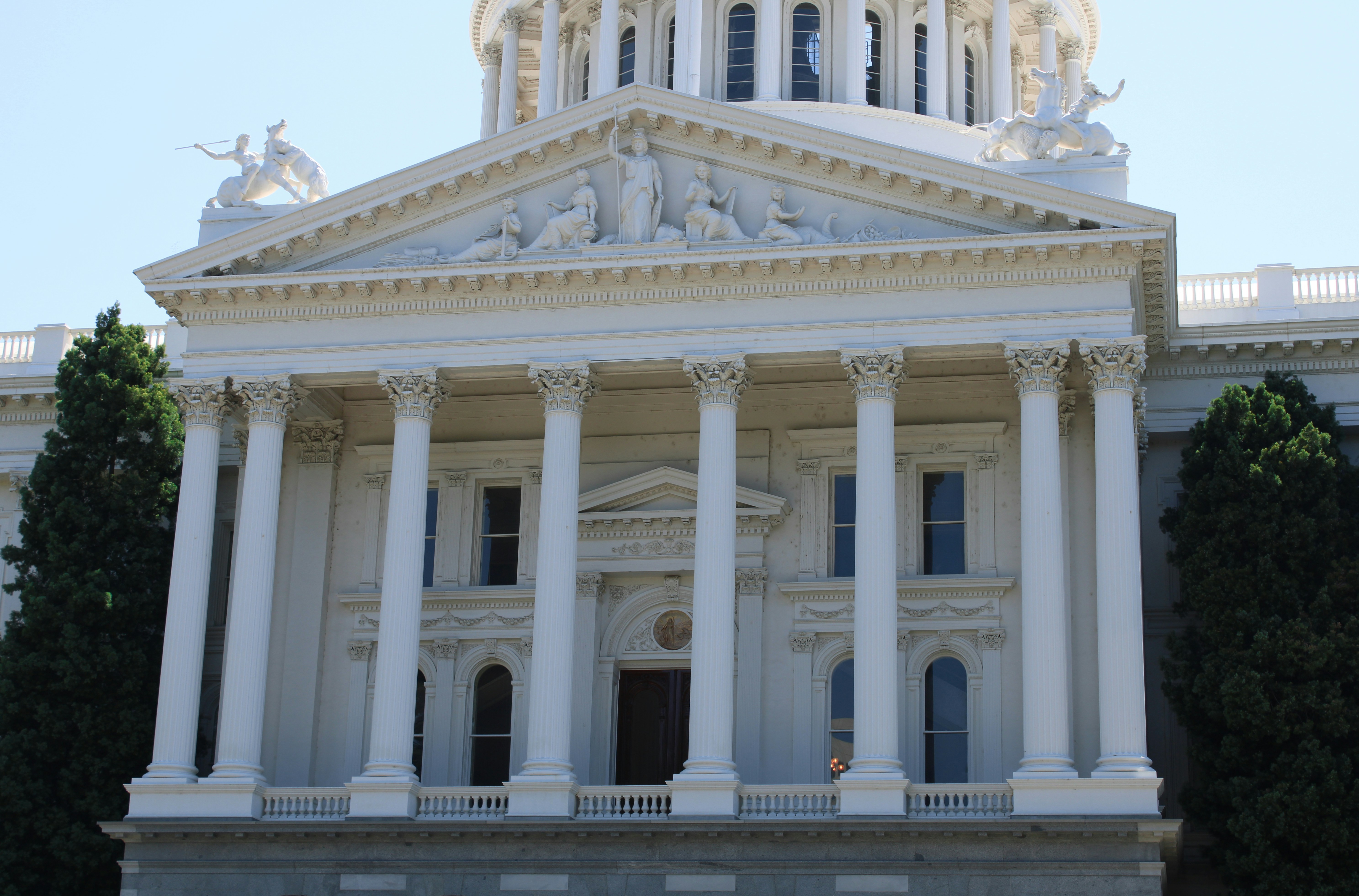 White Neoclassical Building with Dome and Columns A bright white neoclassical building with columned front and partial dome visible, framed by trees and set under clear blue sky. Captured in strong natural light. California Capitol in Sacramento. | The california state capitol building is pictured.