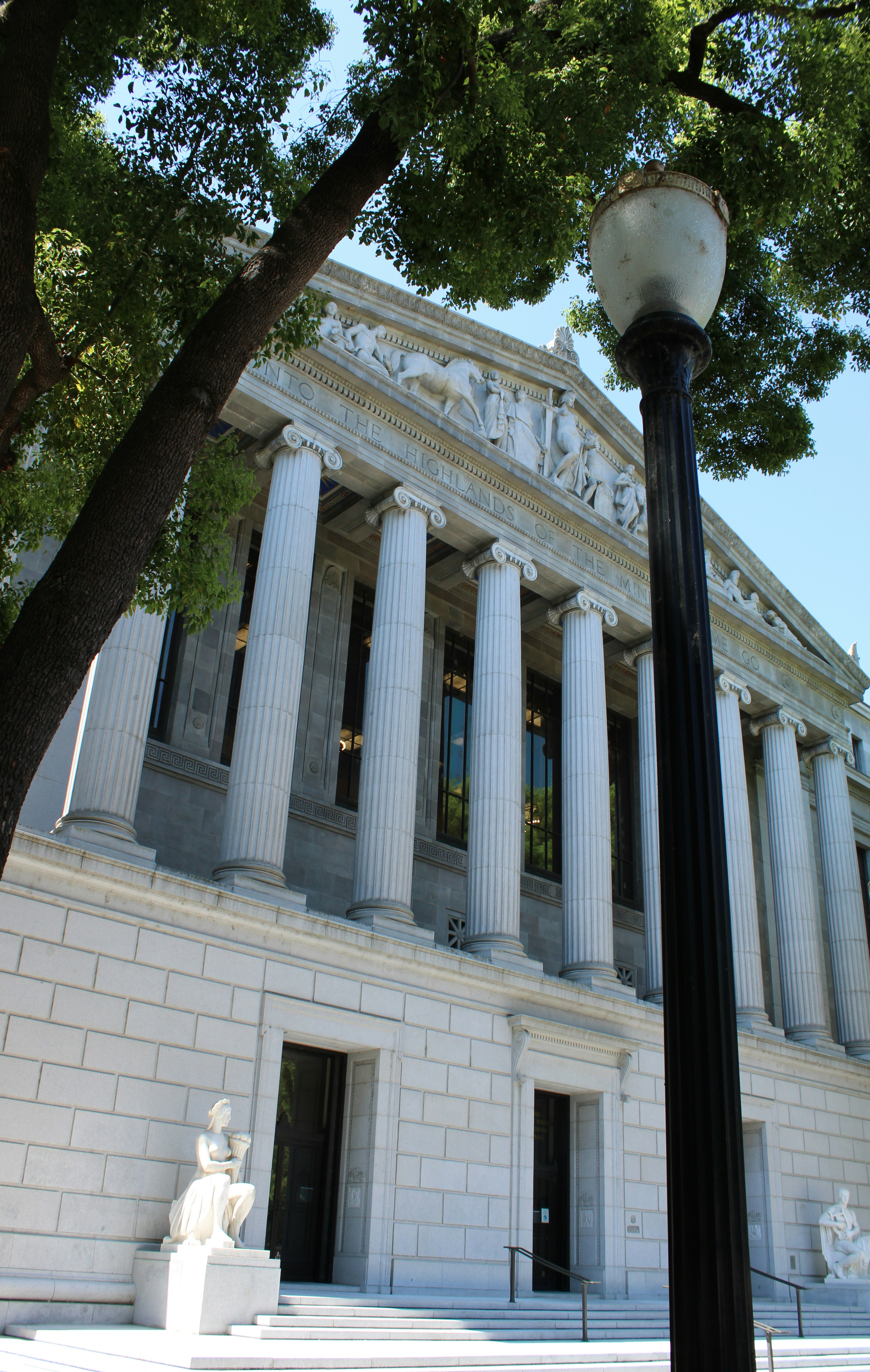 California Capitol Building with Columned Facade and Lamp Post A white columned neoclassical building with a streetlamp in the foreground and a statue at the entrance. Trees frame the scene under bright midday sun. | A classical building with columns and a lamppost.