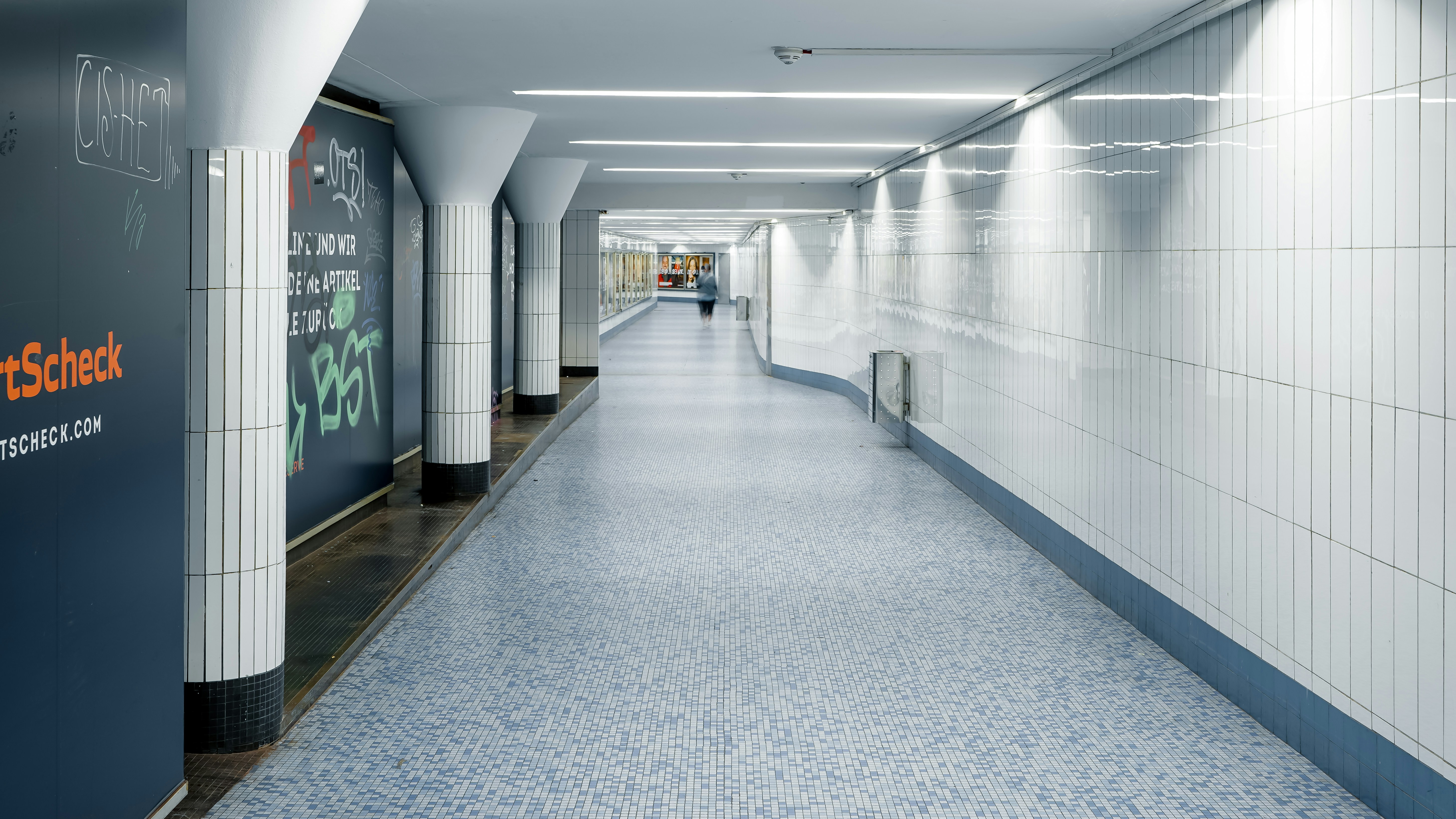 An empty corridor in a modern urban space, featuring tiled walls and floor, illuminated by soft overhead lighting. Graffiti art adds a touch of color to the otherwise minimalist design.