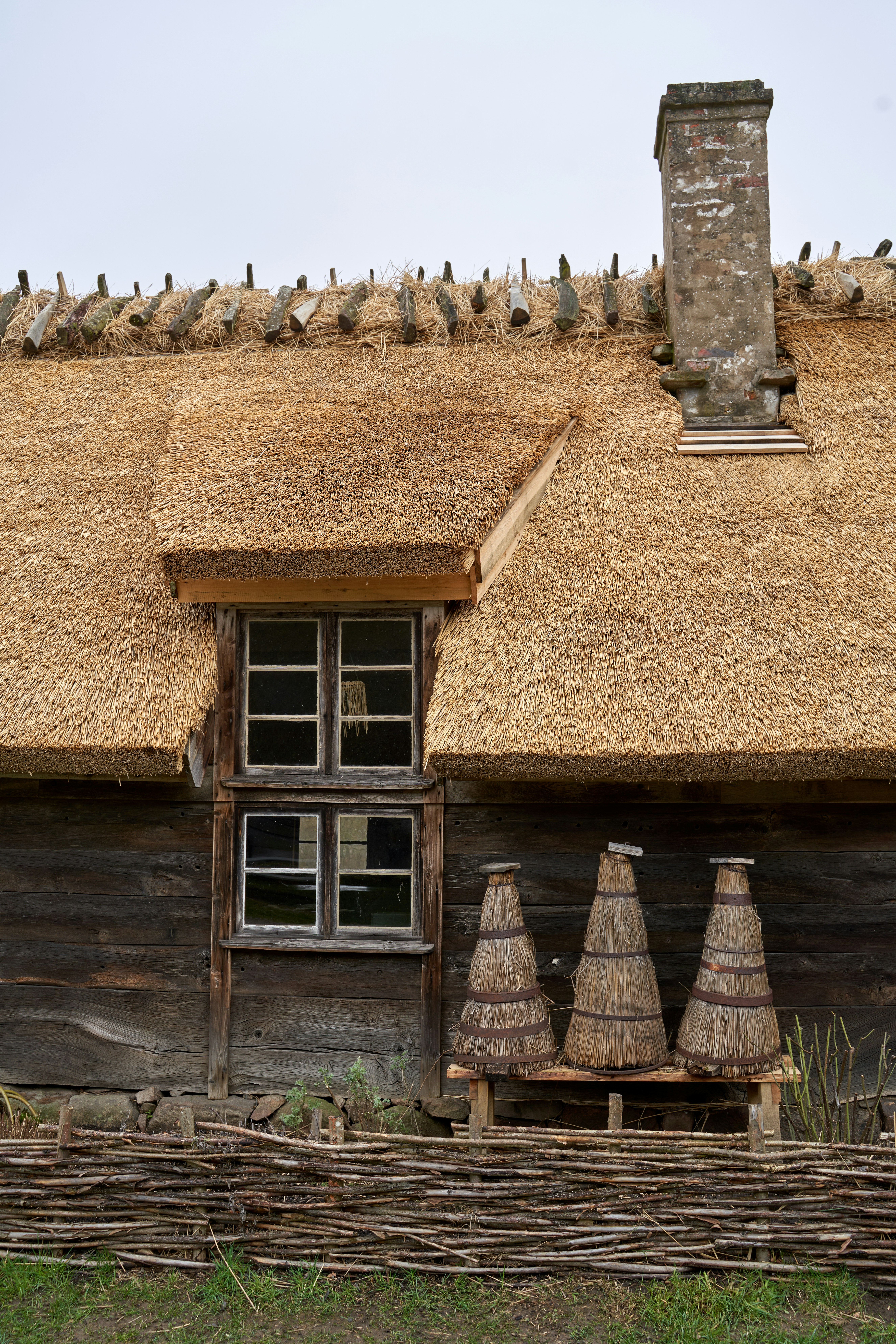 Old timber farm building with reed roof, chimney and wicker wooden fence in Fredriksdal Open-Air Museum, Helsingborg, Sweden | Old cottage with a thatched roof and beehives.