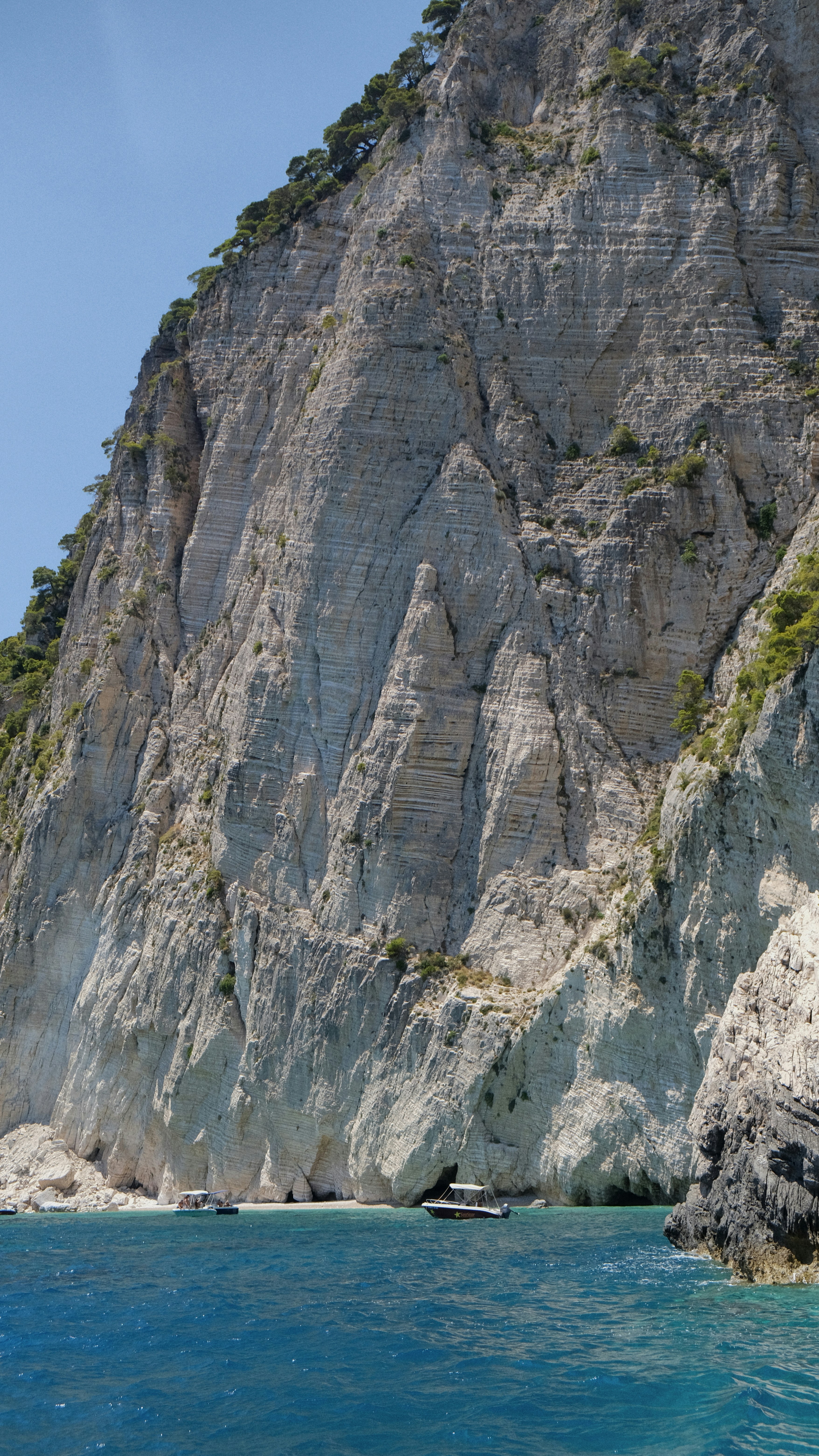 Majestic cliffs rise dramatically from the azure waters, dotted with small boats navigating the shoreline. The rugged textures of the rock formations tell a story of time and nature's artistry.