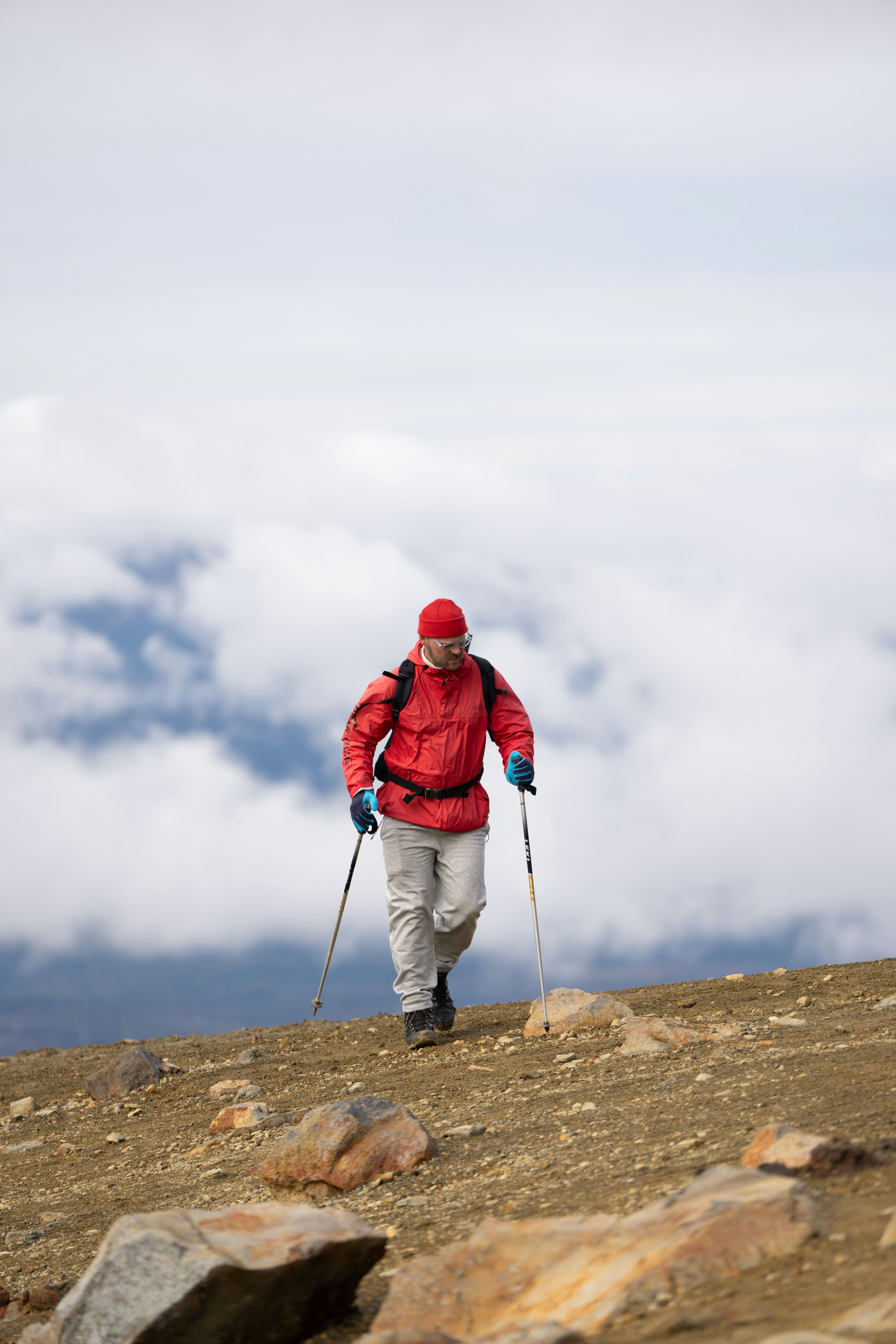 A man in red jacket and hiking gear walks with trekking poles on a rocky mountain trail, surrounded by clouds and high-altitude scenery. | A hiker treks down a hill in cloudy weather.