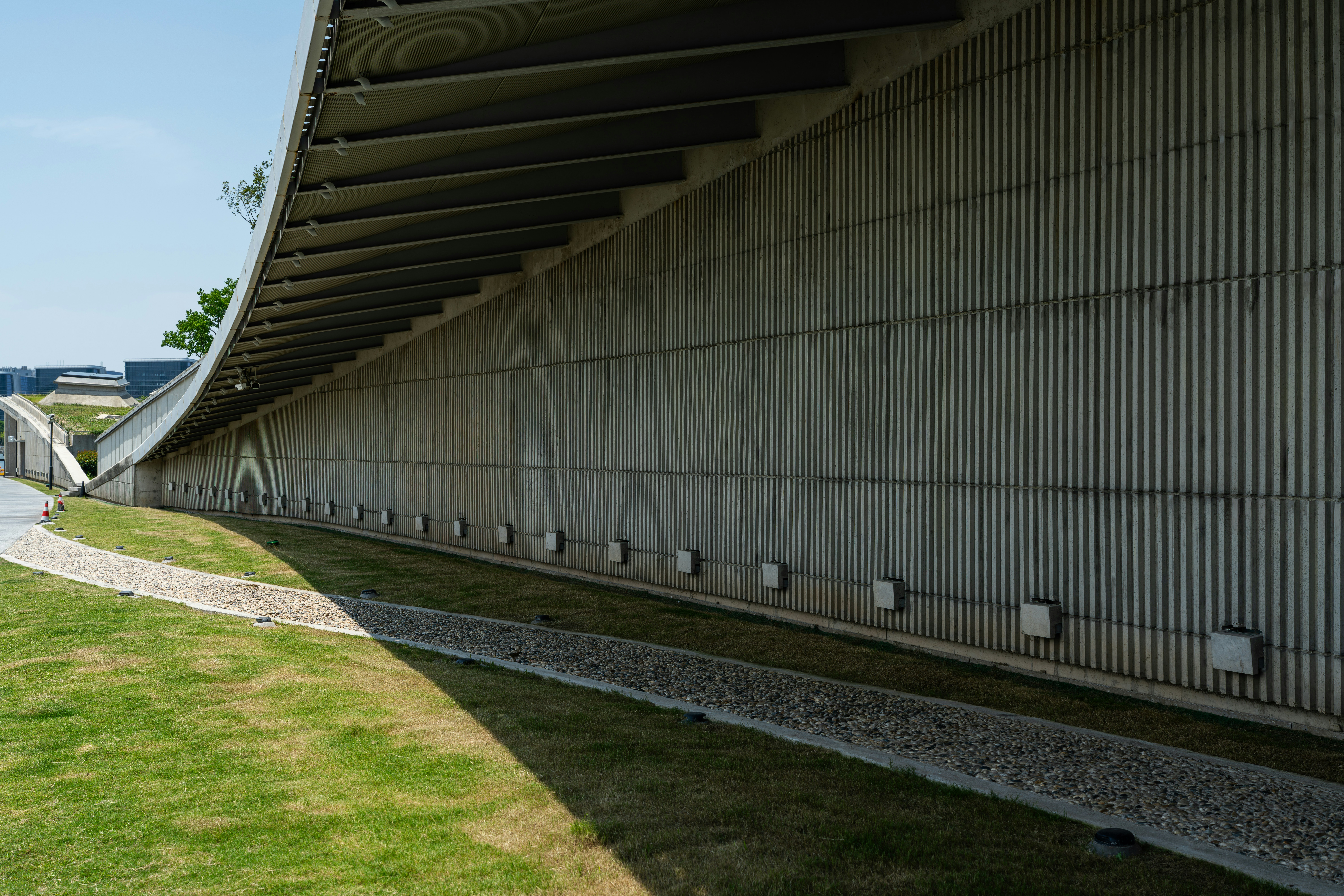 A sweeping concrete wall curves gracefully, contrasting with the lush green grass and gravel path below. The play of light and shadow adds depth to the architectural design.