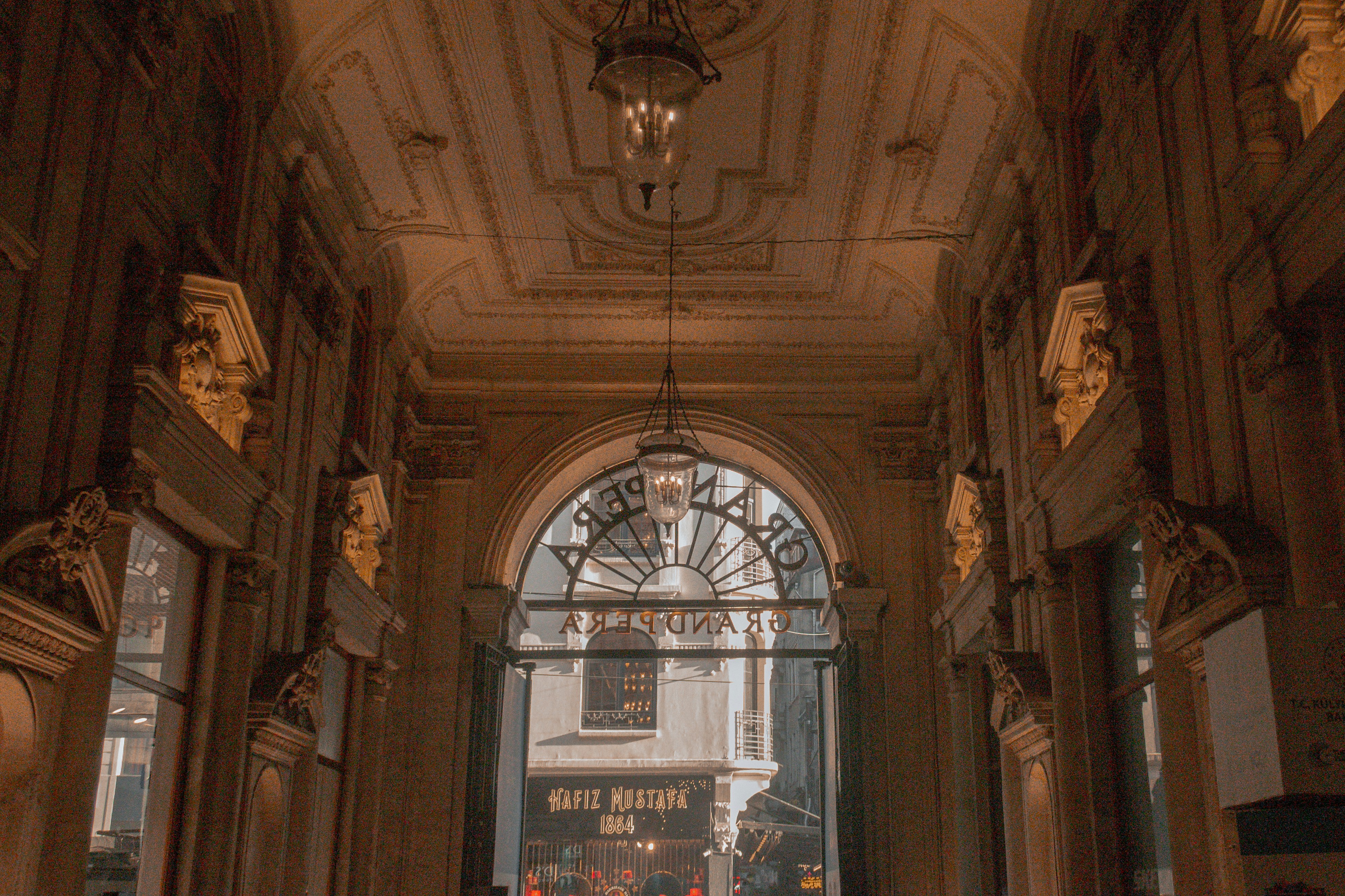 A beautifully lit entrance of a historic passage in Istanbul, featuring intricate neoclassical architectural details. The warm tones, high arched ceiling, and vintage chandeliers create a timeless atmosphere that reflects the elegance of old Istanbul. | A grand archway leads to an exterior view.