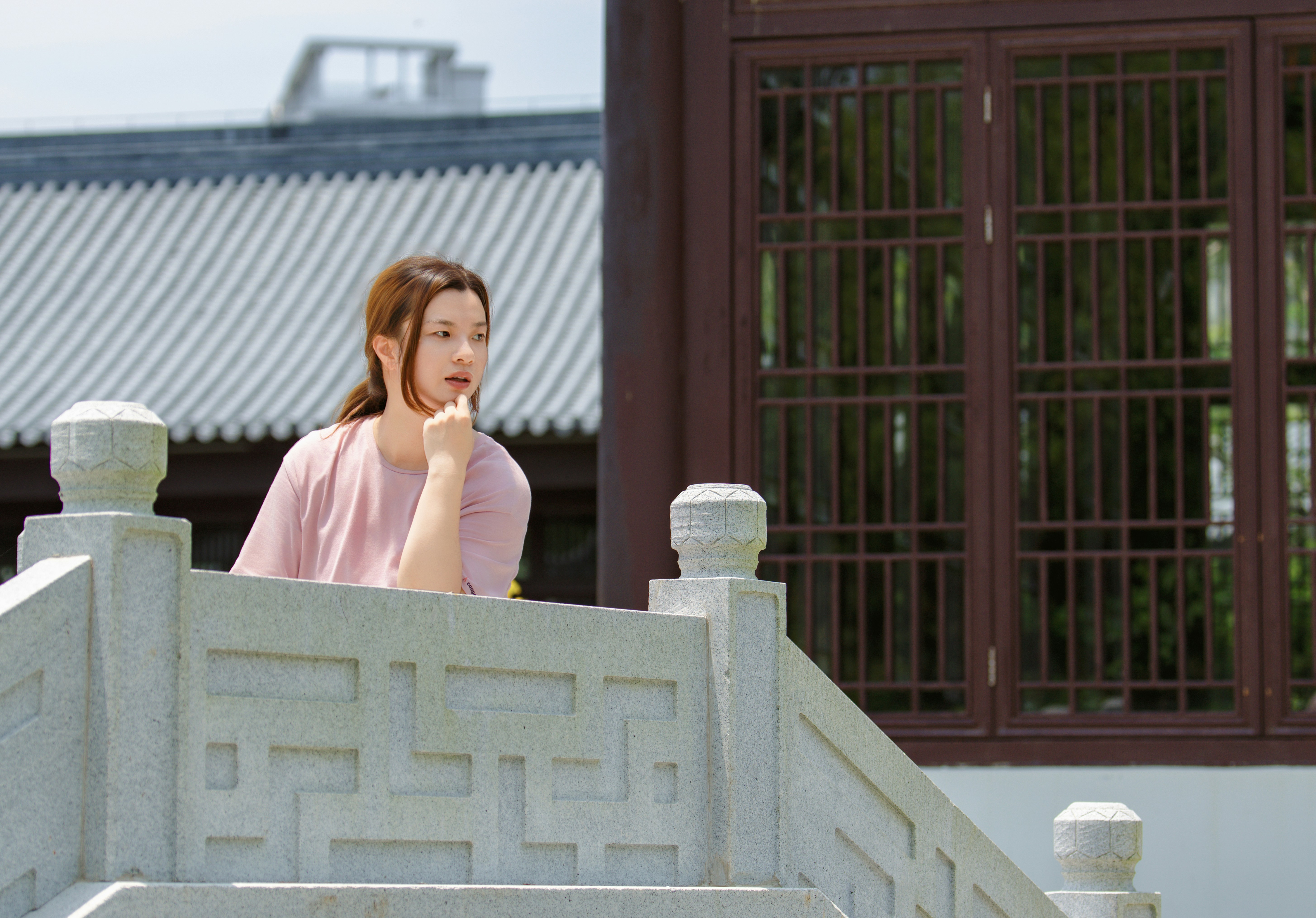 A woman in a pink top thoughtfully poses on a stone bridge, surrounded by traditional architecture and greenery.