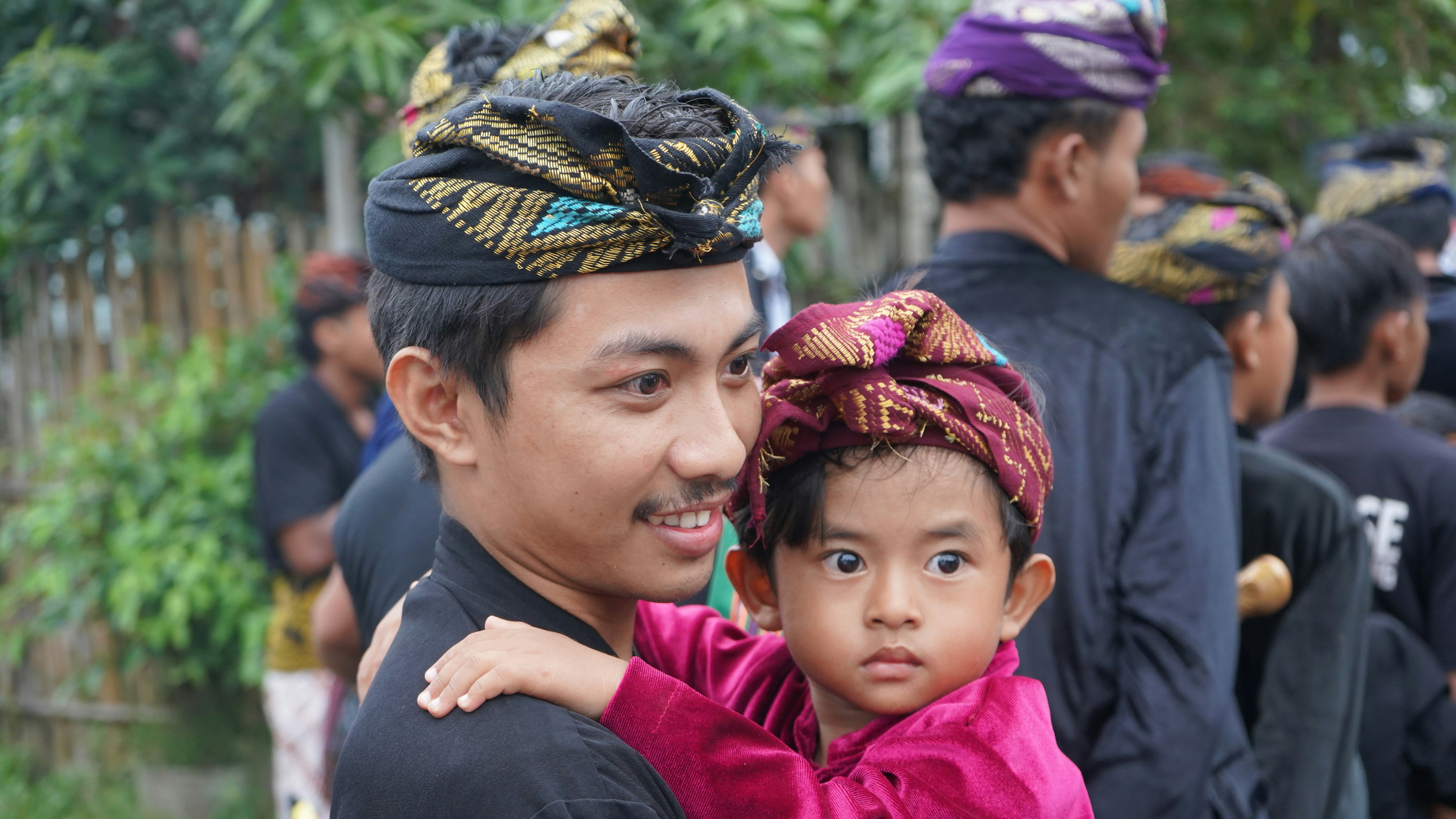 A father and son dressed in traditional Sasak attire share a quiet moment during a village wedding in Lombok, Indonesia. | A man carries a child wearing traditional headdress.